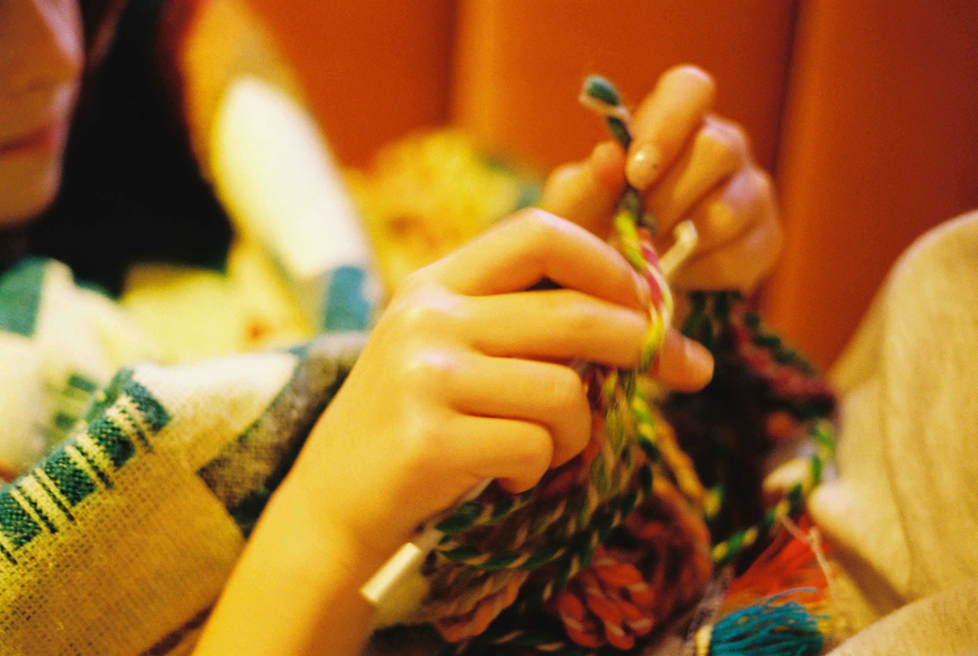 Close-up of hands weaving yarns with colorful threads and a textile in the background; focuses on craftwork and the tactile motion of manipulating fiber. (This is a textile/craft detail, not a logo or illustration.)