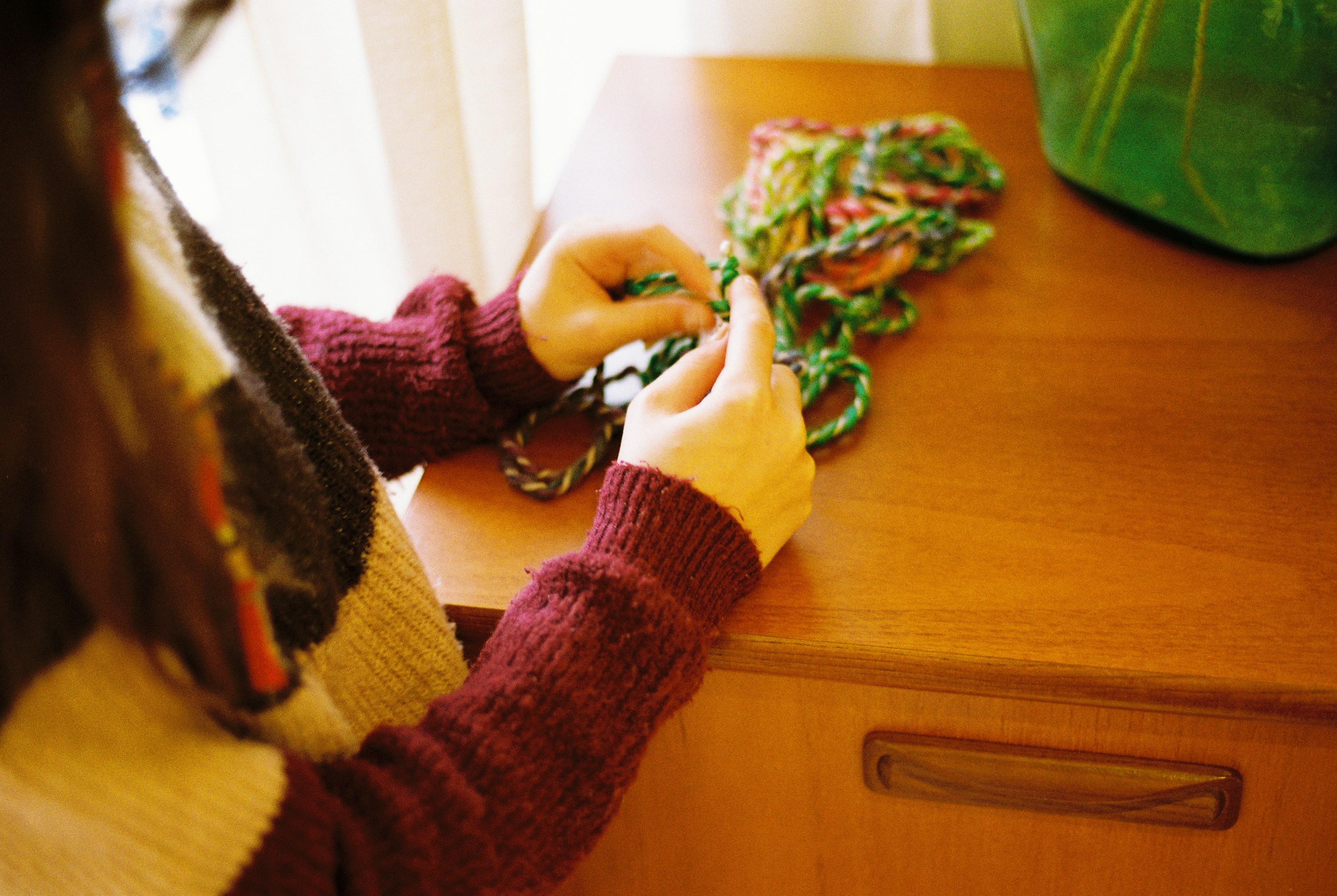 A close view of hands knotting coloured yarn strands into a bracelet on a sunlit wooden desk.