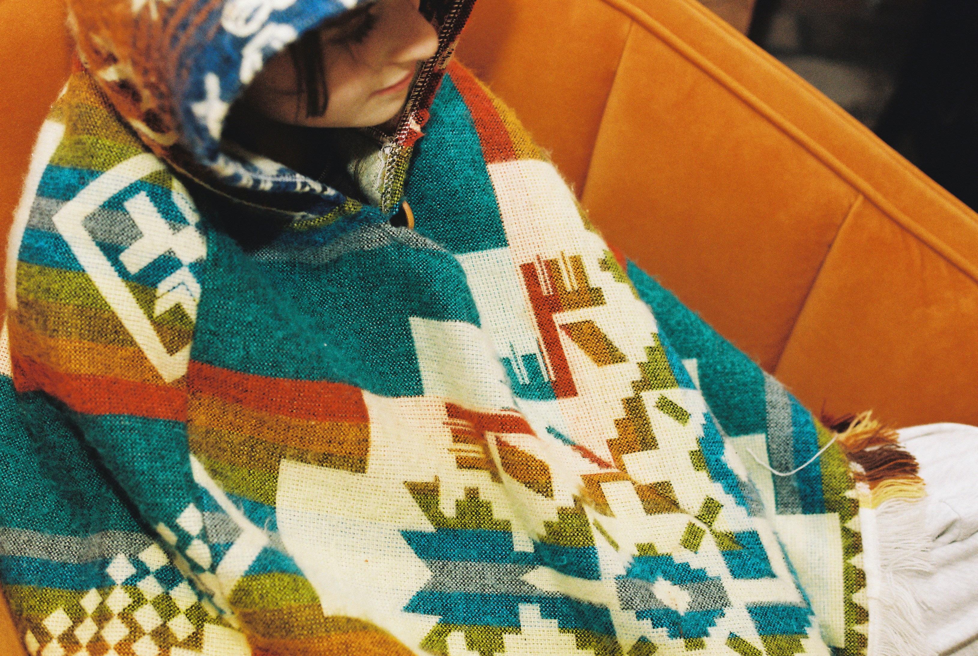 A child in a hooded patterned blanket looks down thoughtfully while curled into a mustard velvet sofa.