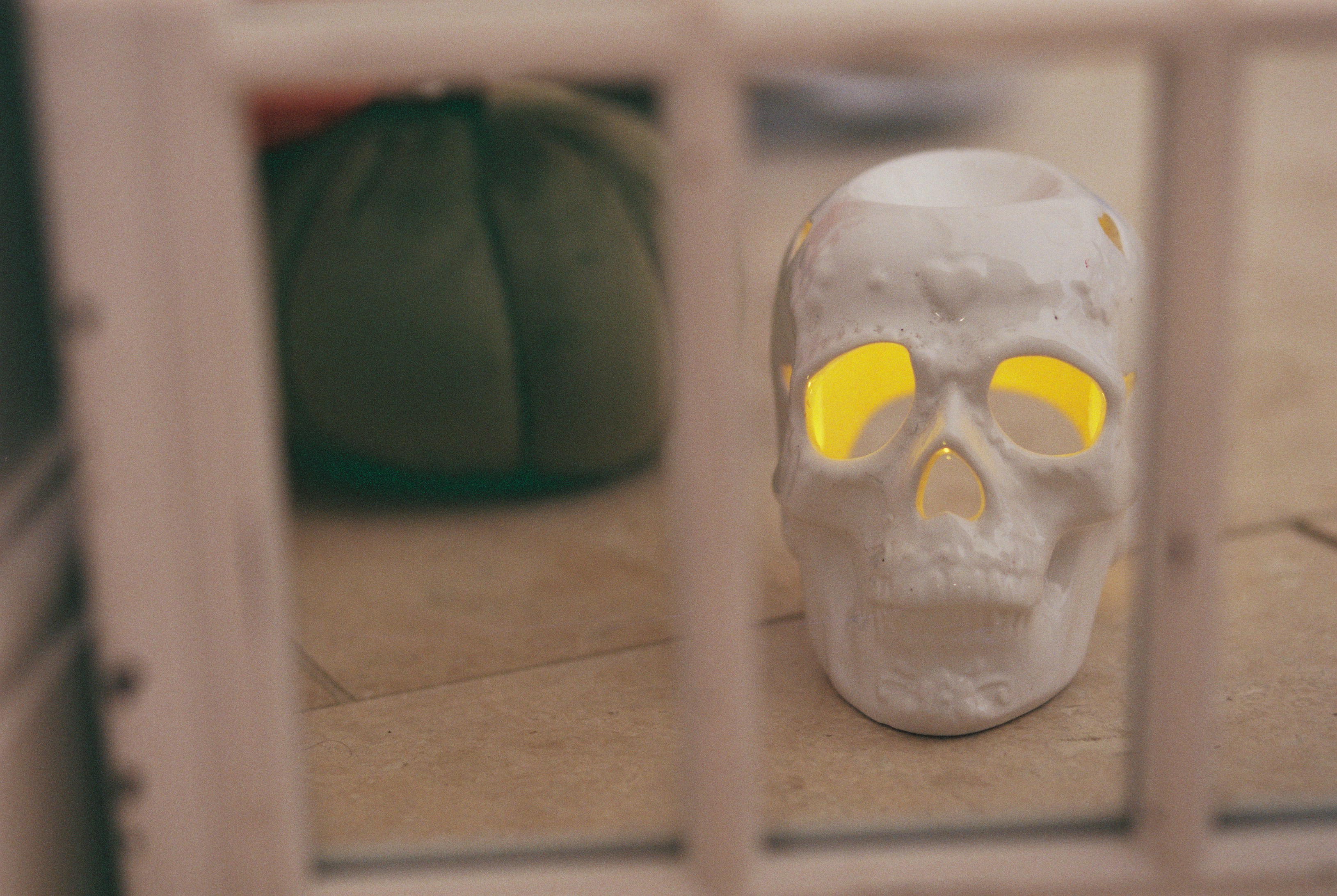 White skull with yellow eye sockets seen through vertical bars, set on a tiled floor with a green cushion in the background. A still-life study of juxtaposed textures and subtle color warmth.
