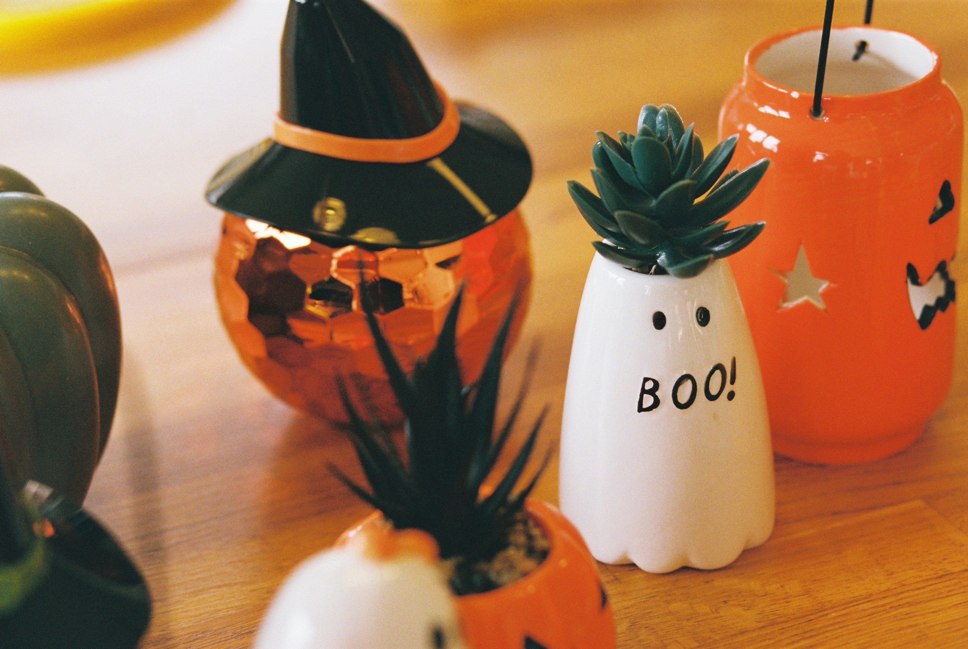 Bright, festive Halloween still-life with small pumpkins and a white Boo! ceramic jar on a wooden table; playful decorations in the background.