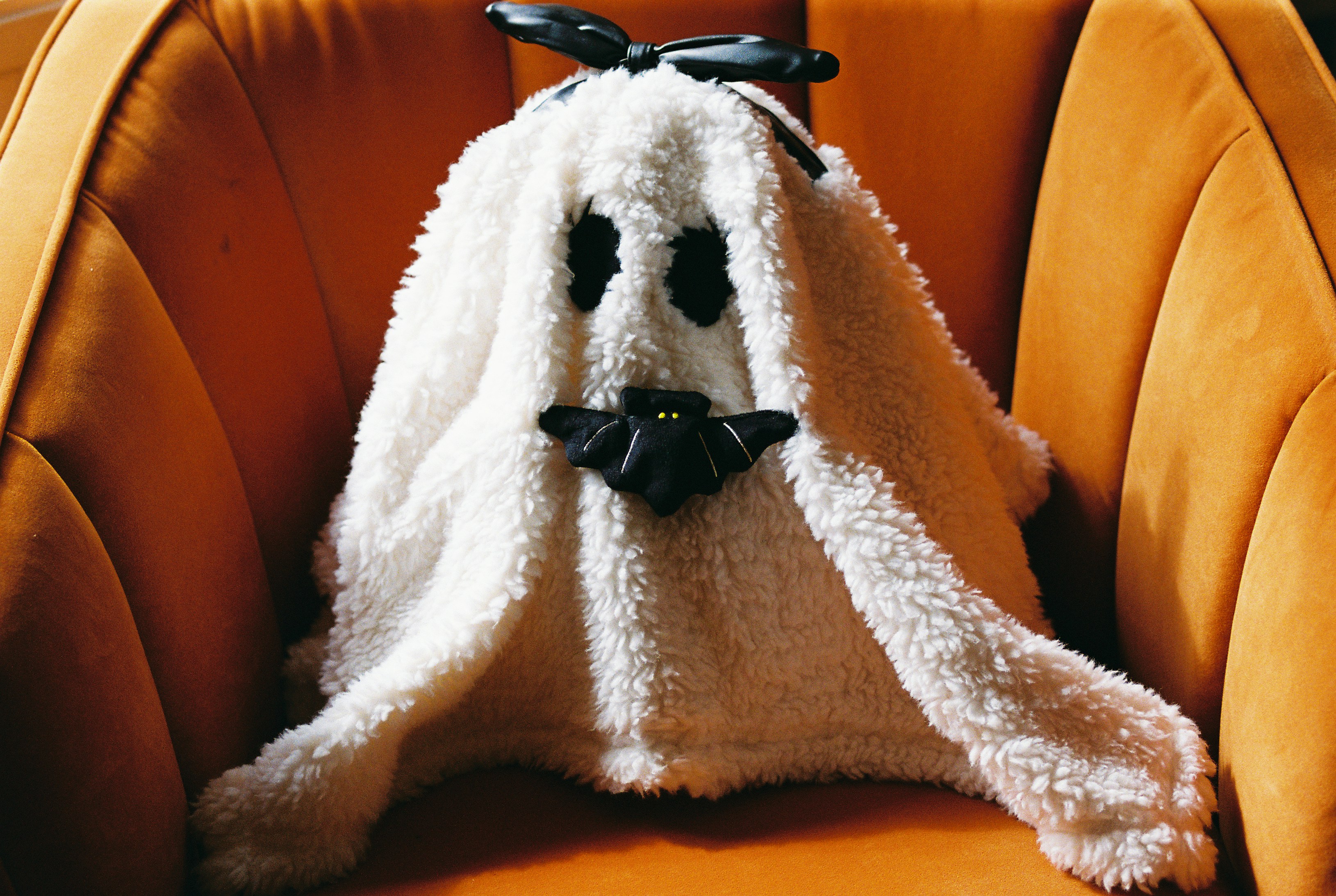 A fluffy sherpa ghost with a bow and bat pin drapes over a mustard velvet armchair in warm light.