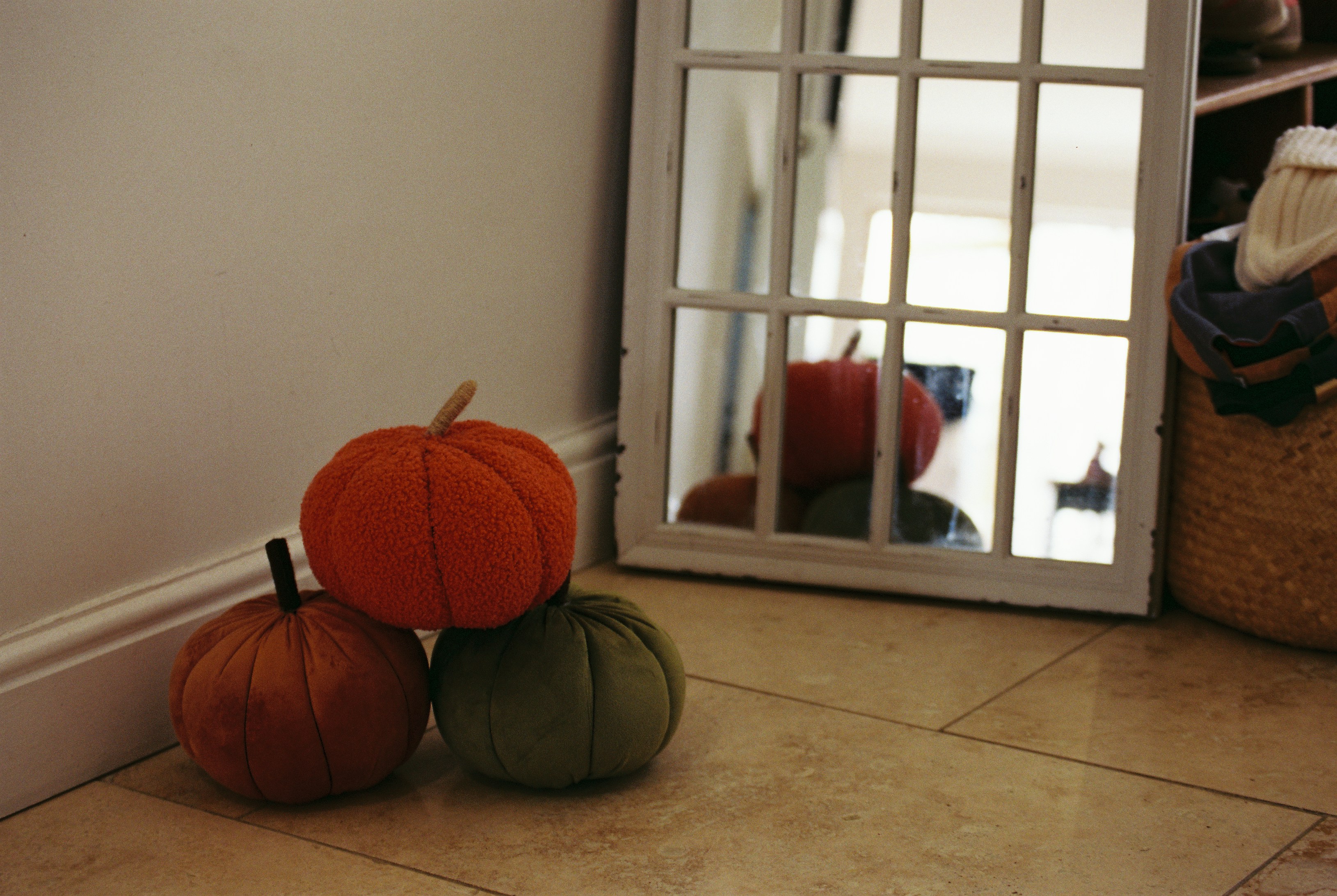 Three pumpkins sit on a tiled floor beside a doorway, with a mirror reflecting the interior. A woven basket and a soft, cozy scene frame the warm, autumnal still life.