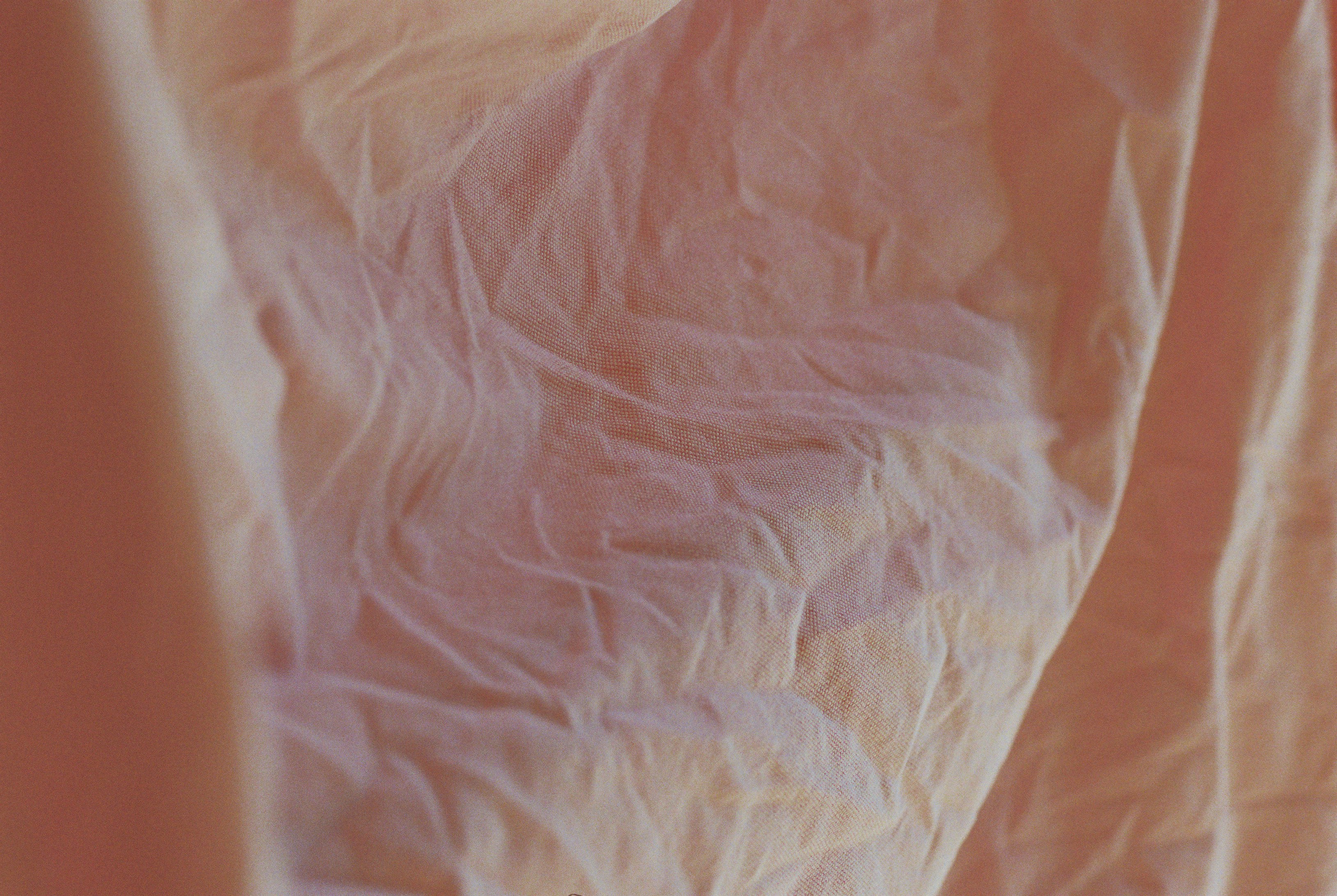 Textured fabric close-up with warm, peach-toned lighting showing creases and folds in the material. Photography focuses on the tactile surface and subtle color gradations; no discernible subject beyond the fabric.