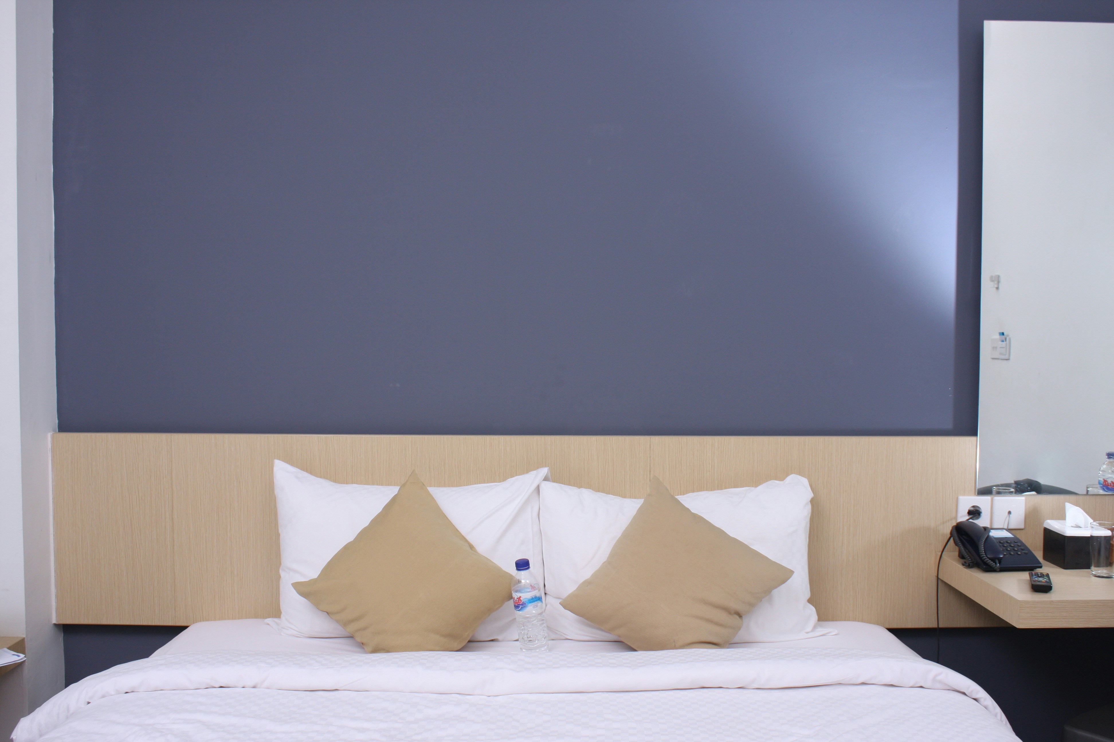 A tidy hotel bed with white linens and three decorative pillows sits against a blue accent wall, flanked by a nightstand and practical desk setup.
