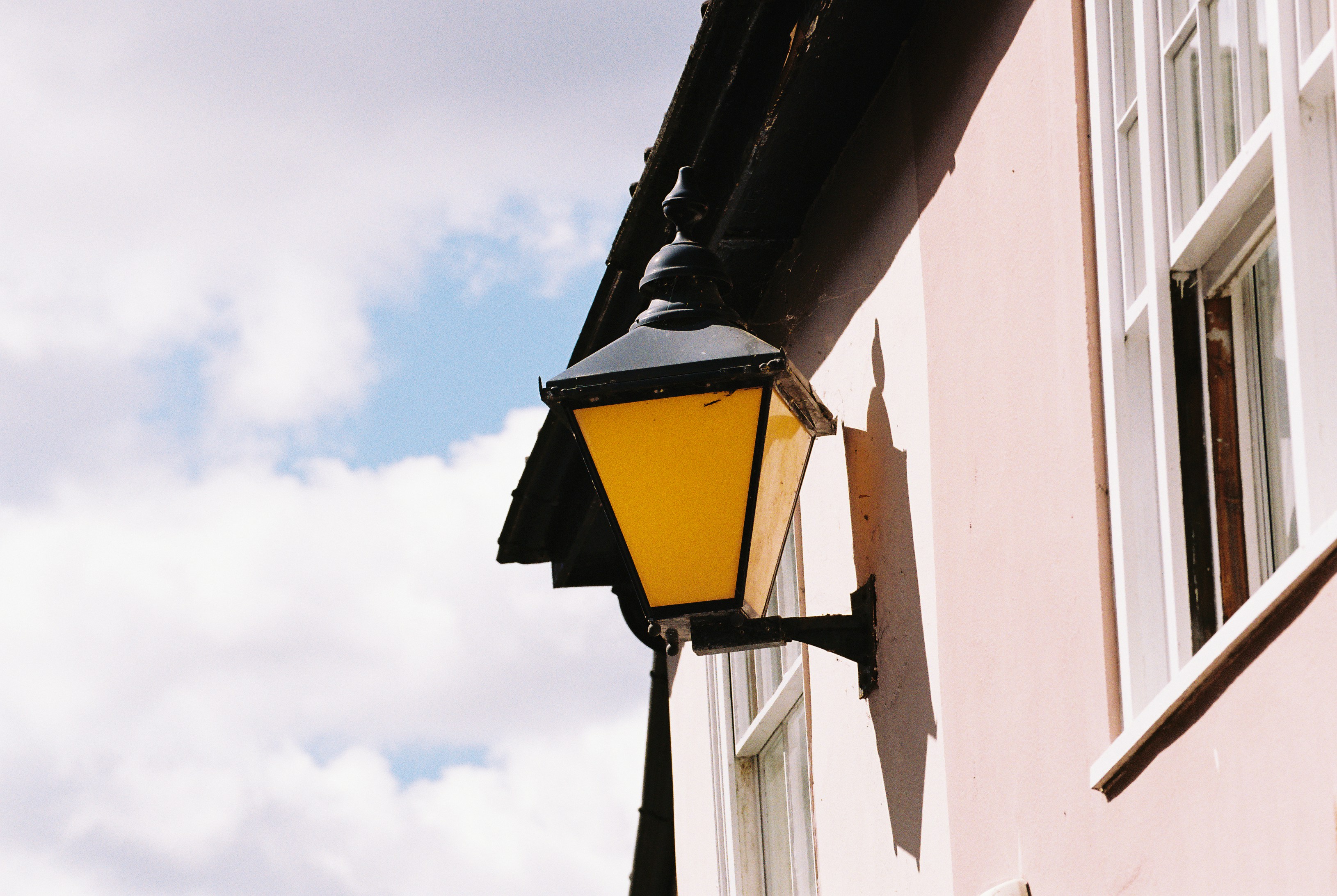 A black street lantern with an amber glass pane juts from a pink rendered wall under a bright cloud-scattered sky.