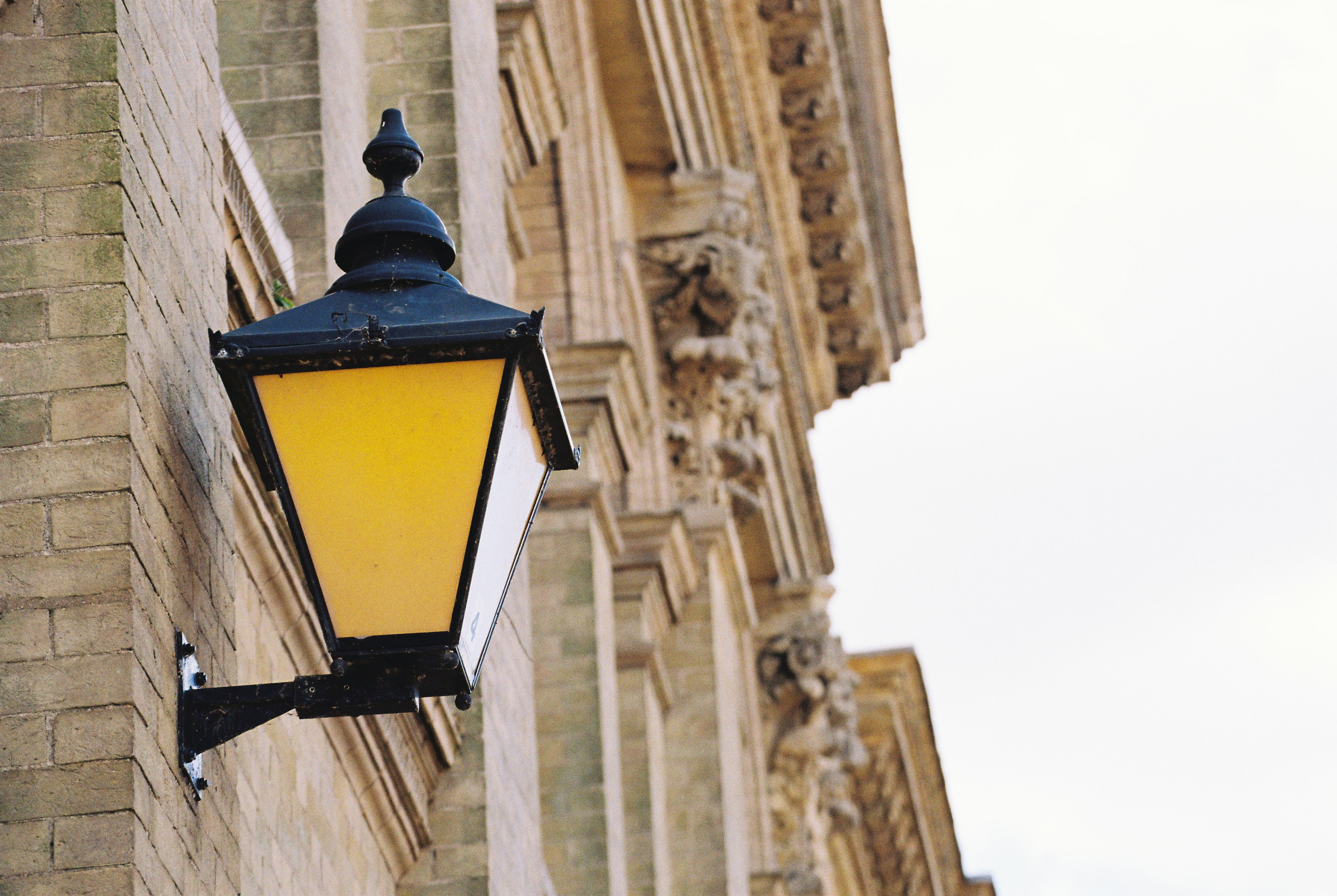 A warm, yellow street lantern projects from a brick building with ornate stonework along a light sky; the scene emphasizes classic architectural detail. (Logotype/indoor not present).