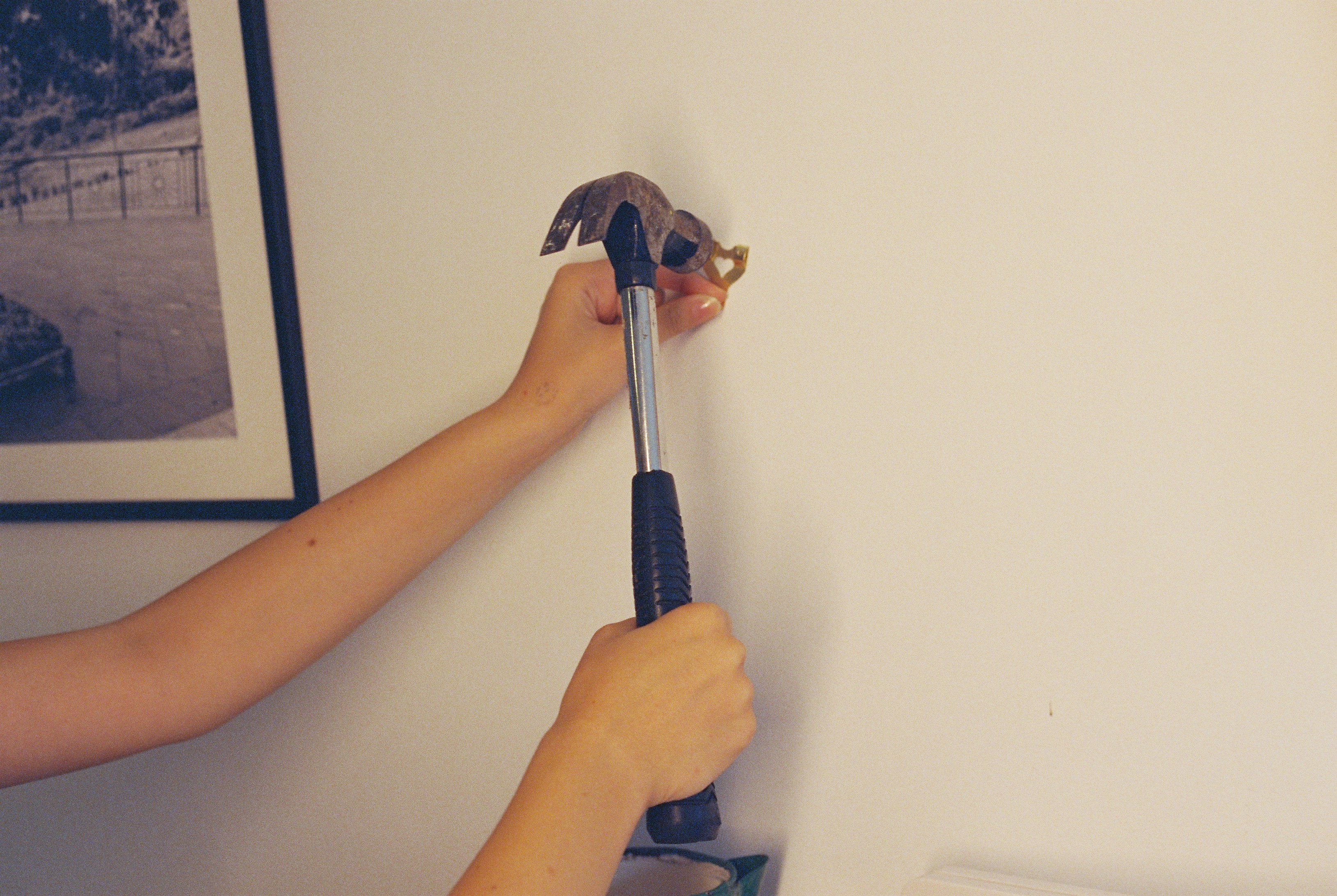 Person driving a hammer into a wall, mounted on the wall is a framed photo; hand and hammer near the center, wall and a small tool chest visible in the lower area.
