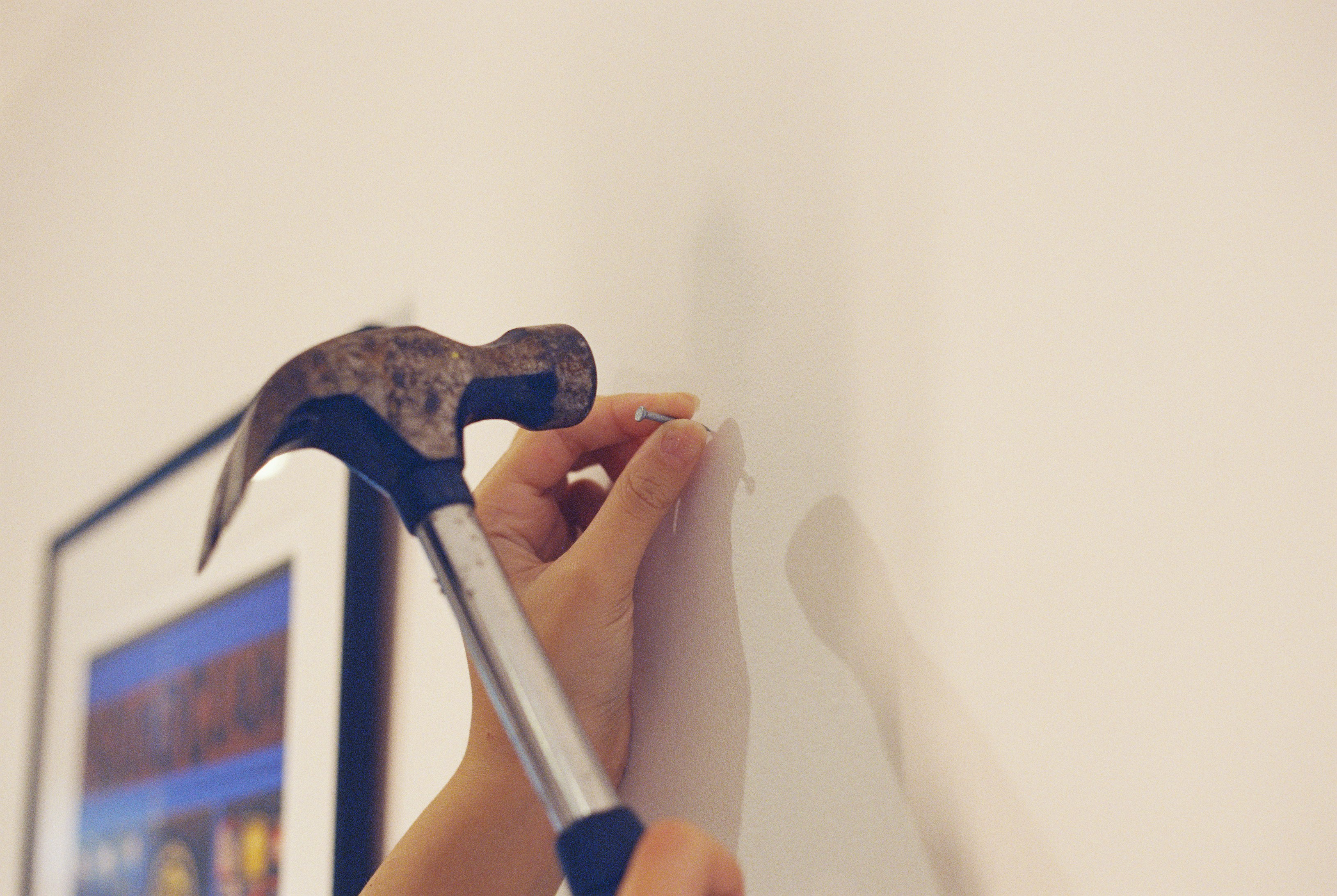 A hand holding a hammer marks a wall beside a framed picture, captured in warm, soft light.