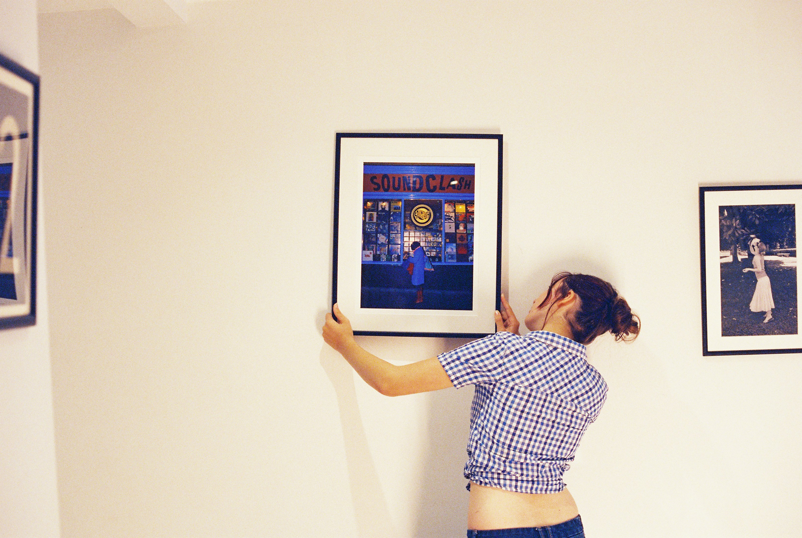A person in a checkered top adjusts a framed artwork on a pale wall, with additional frames visible on either side, capturing a casual museum-like scene.