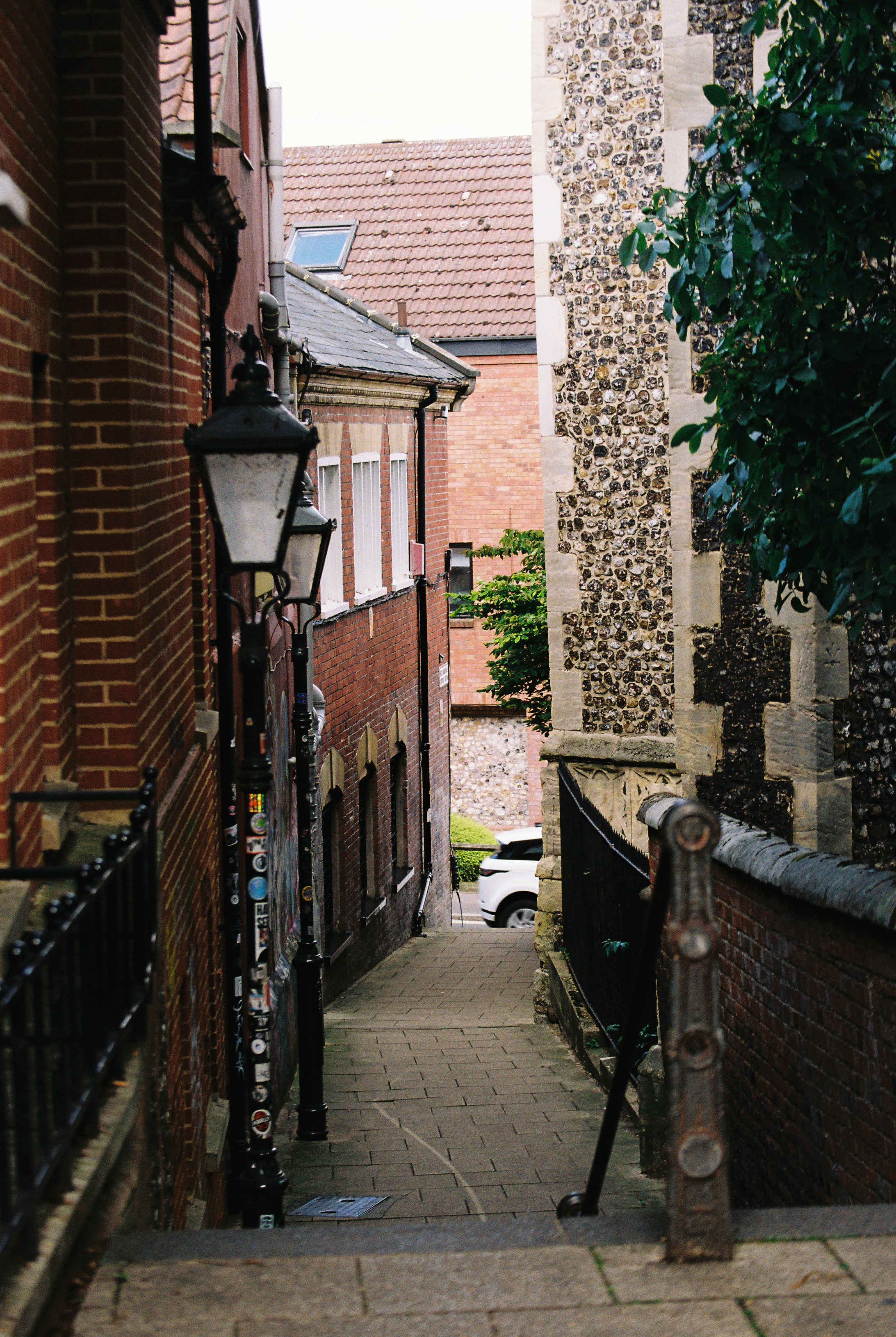 Worn steps drop into a quiet backstreet where a sticker-covered lamp post stands against brick and flint walls.