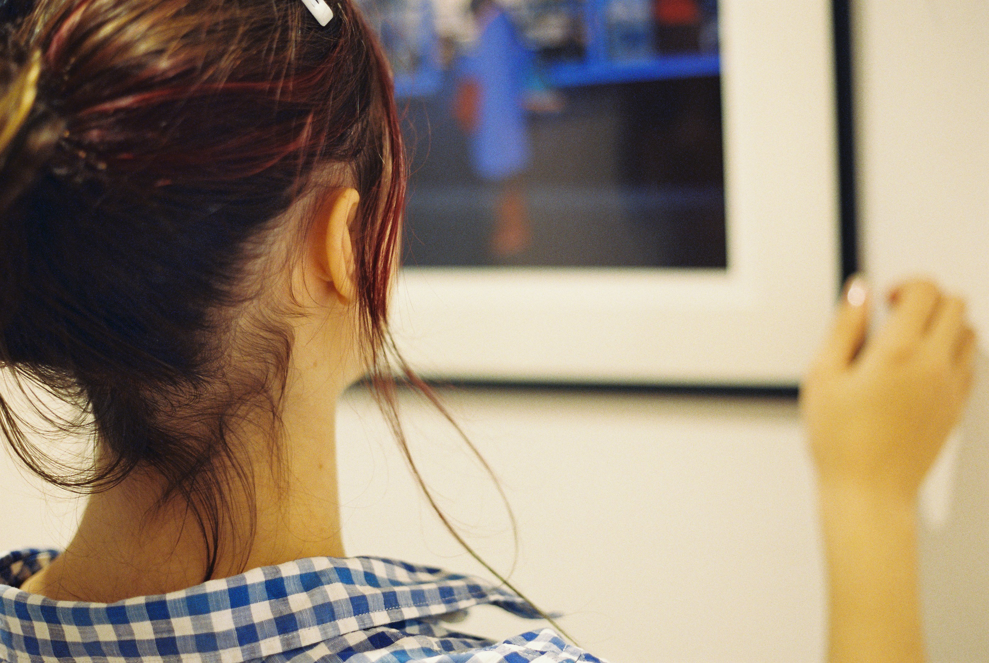 Person viewed from behind adjusting a wall-mounted monitor; blue-checkered shirt and hair tied back, framed by the edge of a wall and framed display.