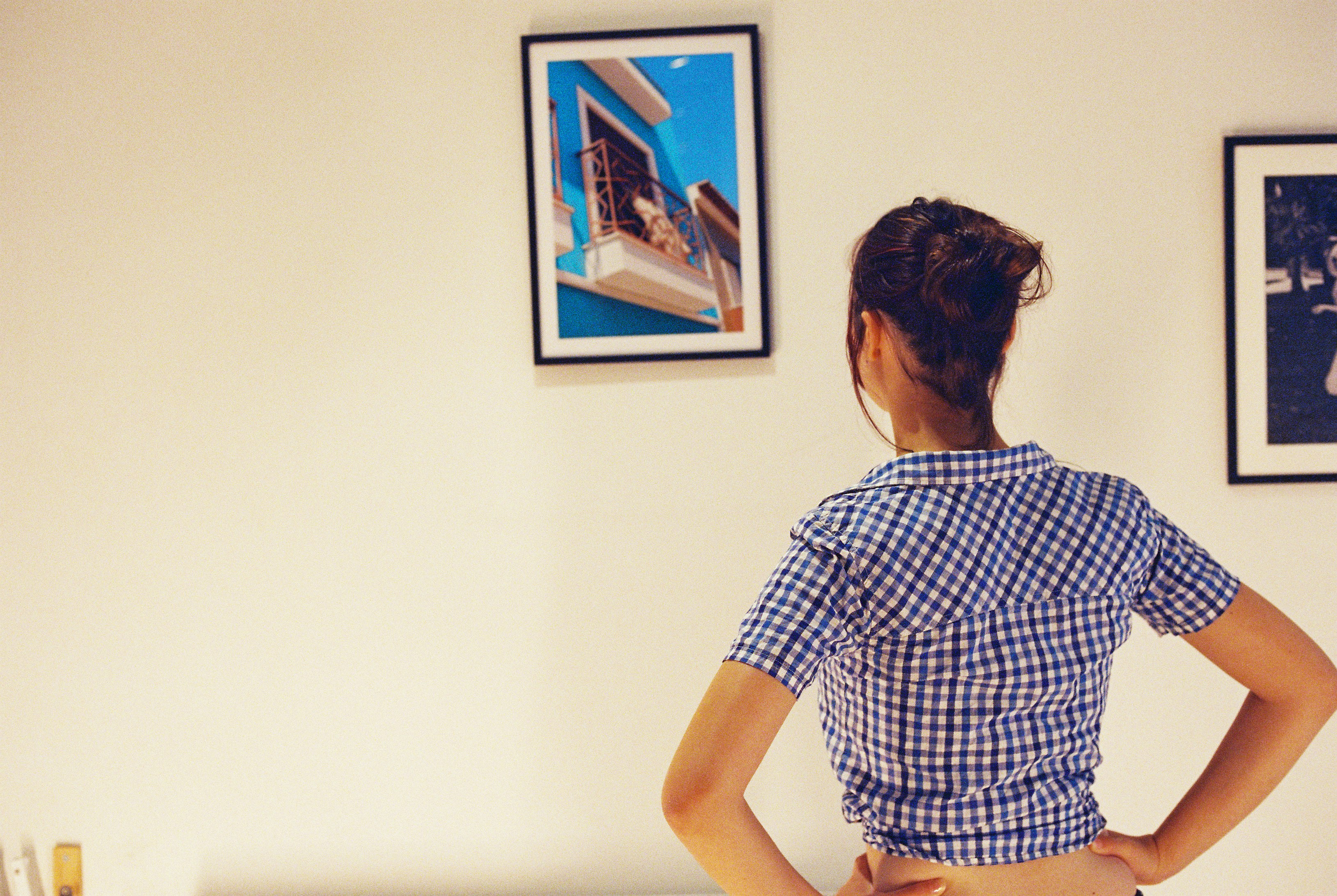 A person stands with hands on hips facing framed photos on a light wall, emphasizing a casual interior portrait with a checkered top.