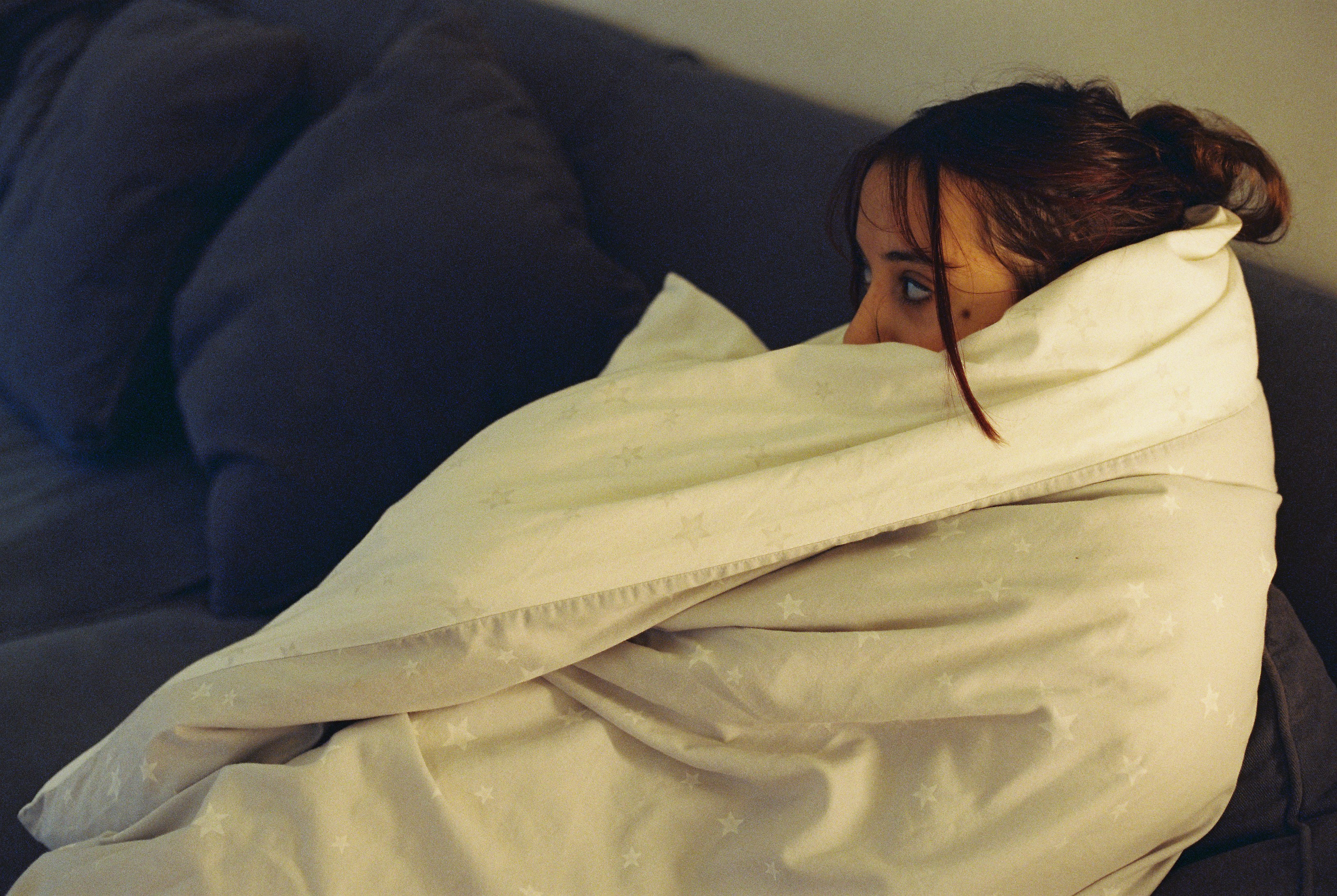 A woman sits wrapped in a pale blanket on a sofa, her face peeking from under the coverings as she gazes off to the side. The scene emphasizes coziness and domestic stillness, with soft lighting and muted colors. (Photographic still).