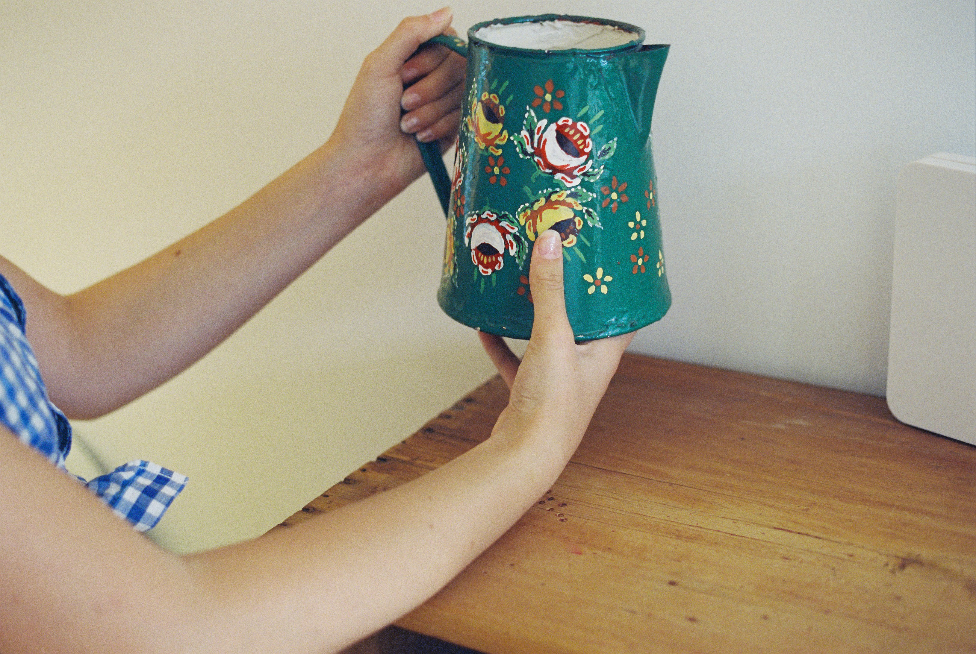 A teal floral pitcher is held by two hands above a wooden table, with a soft indoor light in the background. This image focuses on the object being manipulated, not a posed face; it is a still-life style capture of a kitchen scene.