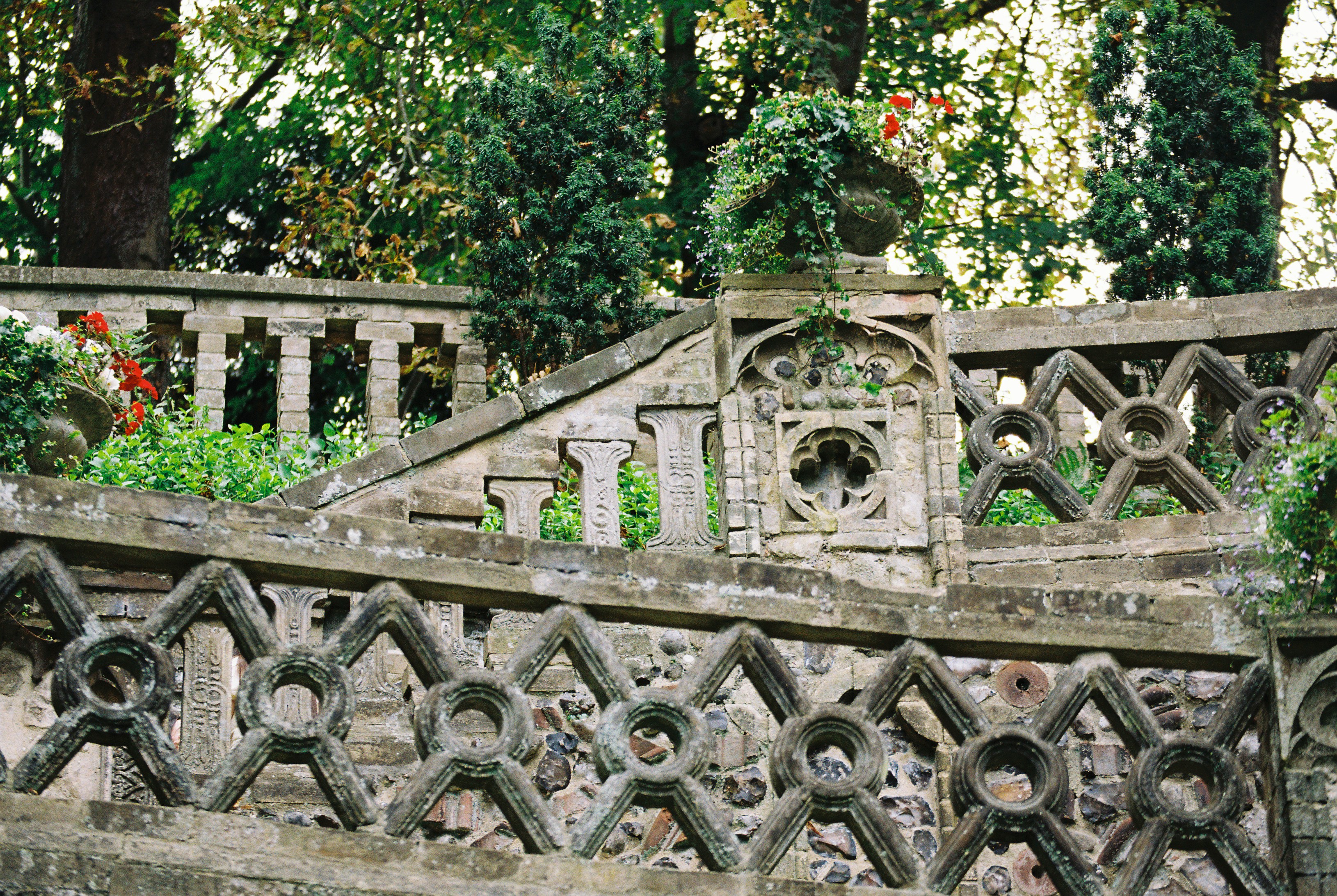 Weathered stone balustrades and ornate railing rise beside a lush garden, with greenery and red flowers framing the architectural details.