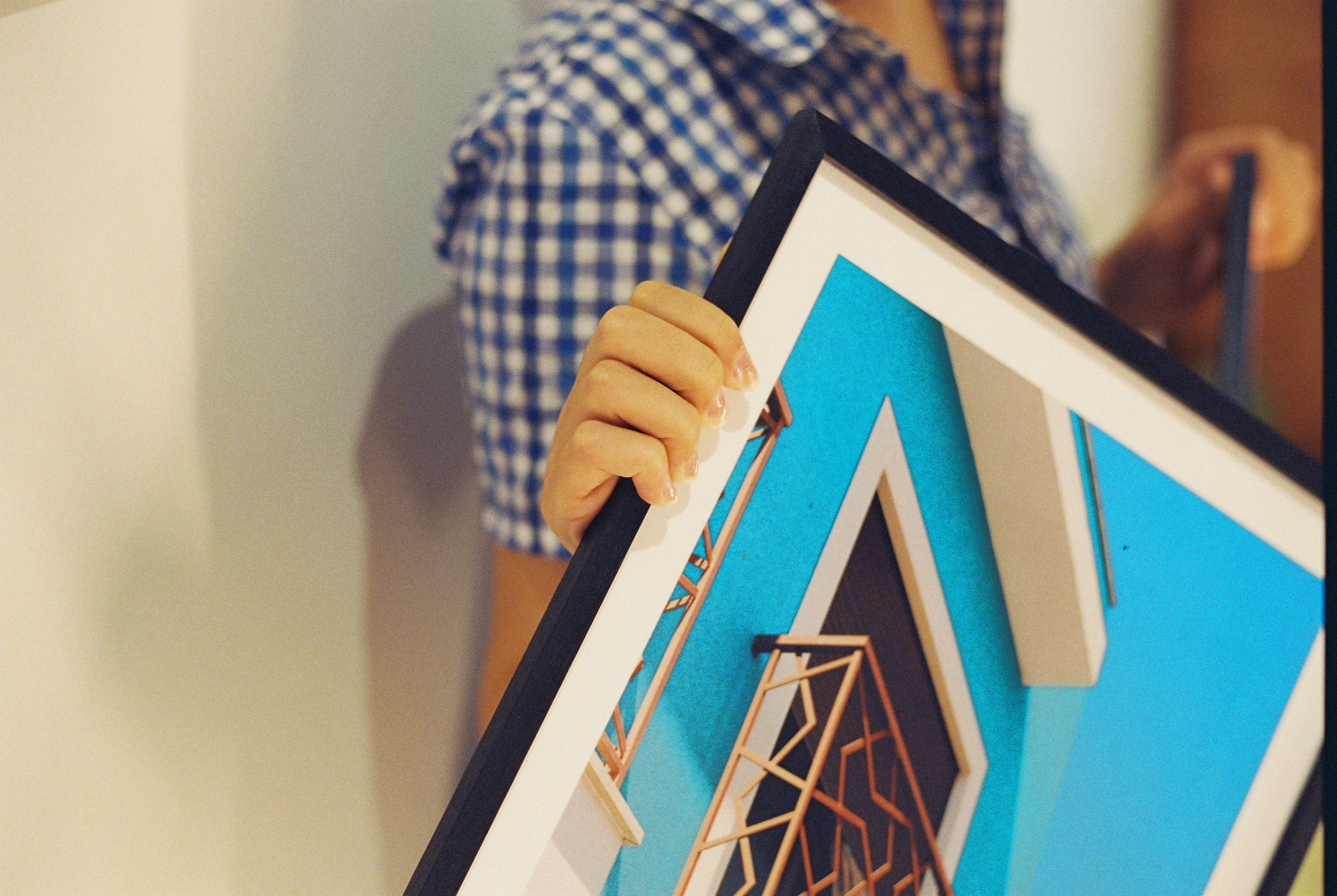A person holds a tilted framed artwork featuring bright blue geometric shapes, inviting a close look at color and lines in a softly lit interior.