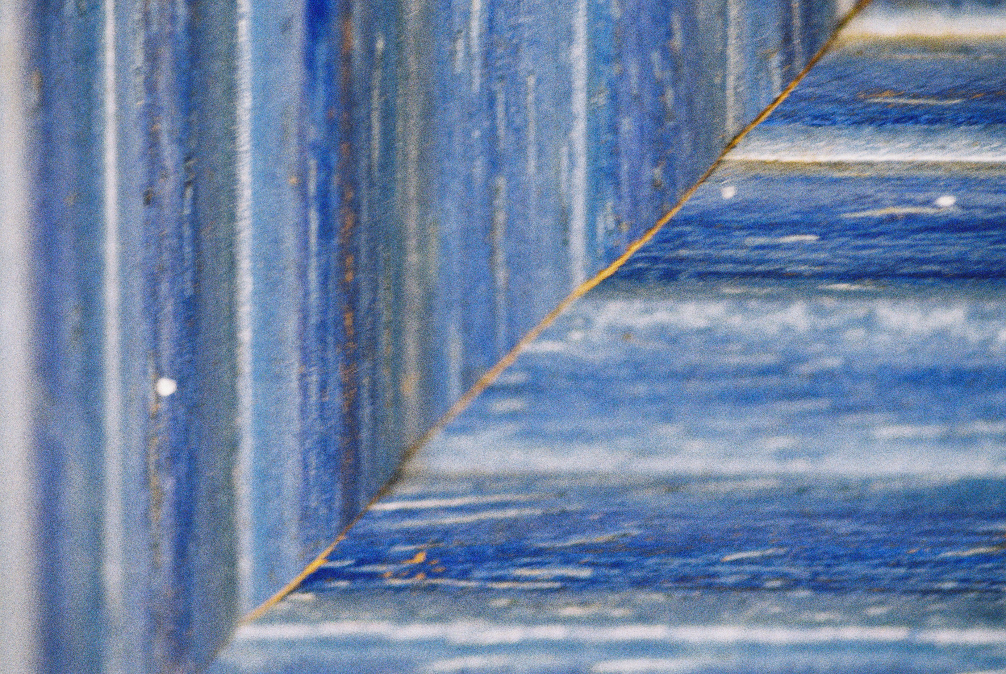 Blue-painted wooden steps forming converging diagonal lines with weathered texture and small white specks; abstract architectural detail without a distinct subject.