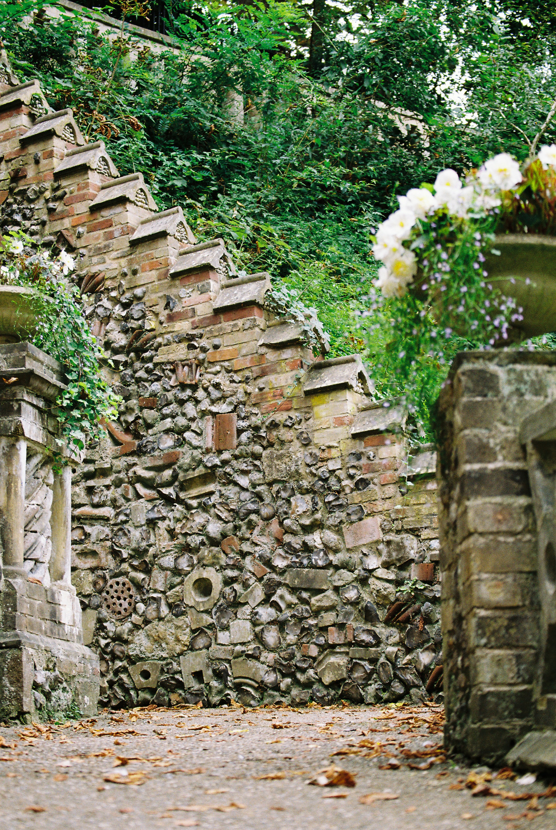 Fallen leaves scatter across gravel beside a stepped wall of flint, brick and shells, with white roses spilling from a stone urn.