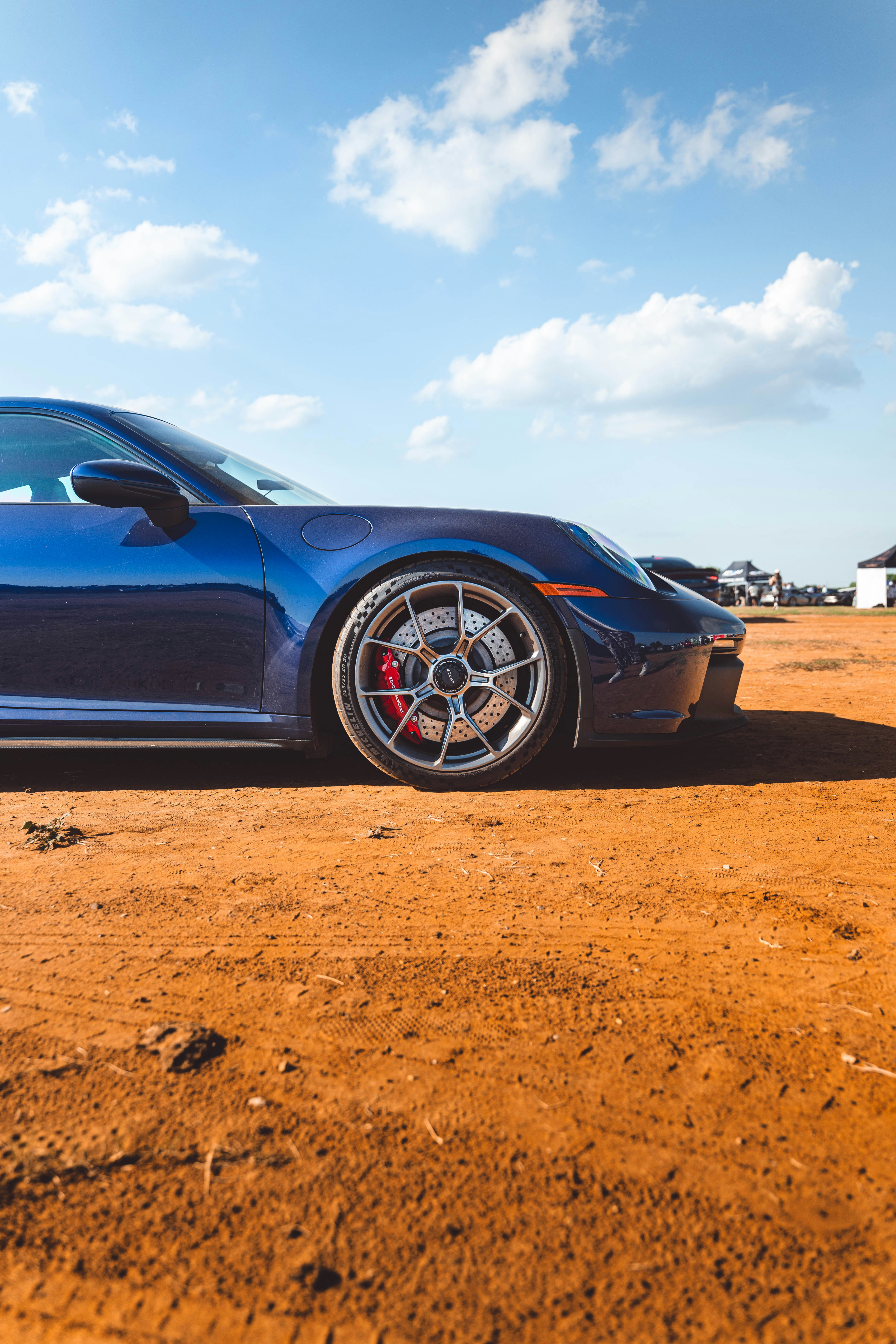 Sleek blue Porsche parked on sandy terrain under a bright blue sky, highlighting the wheel detail and stance in a wide outdoor setting.