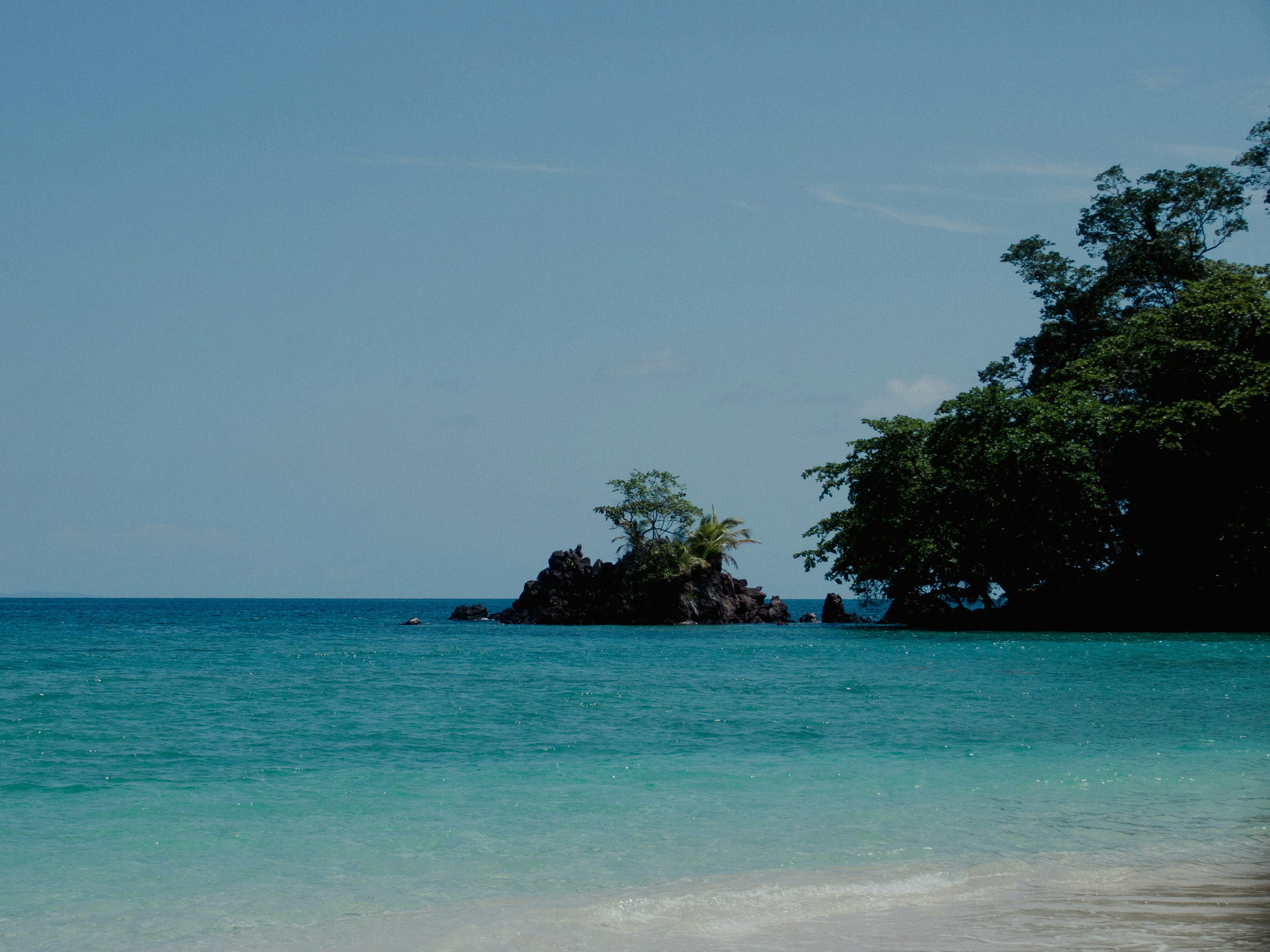 Sandy shoreline with turquoise water and a small rocky island framed by trees in the distance.
