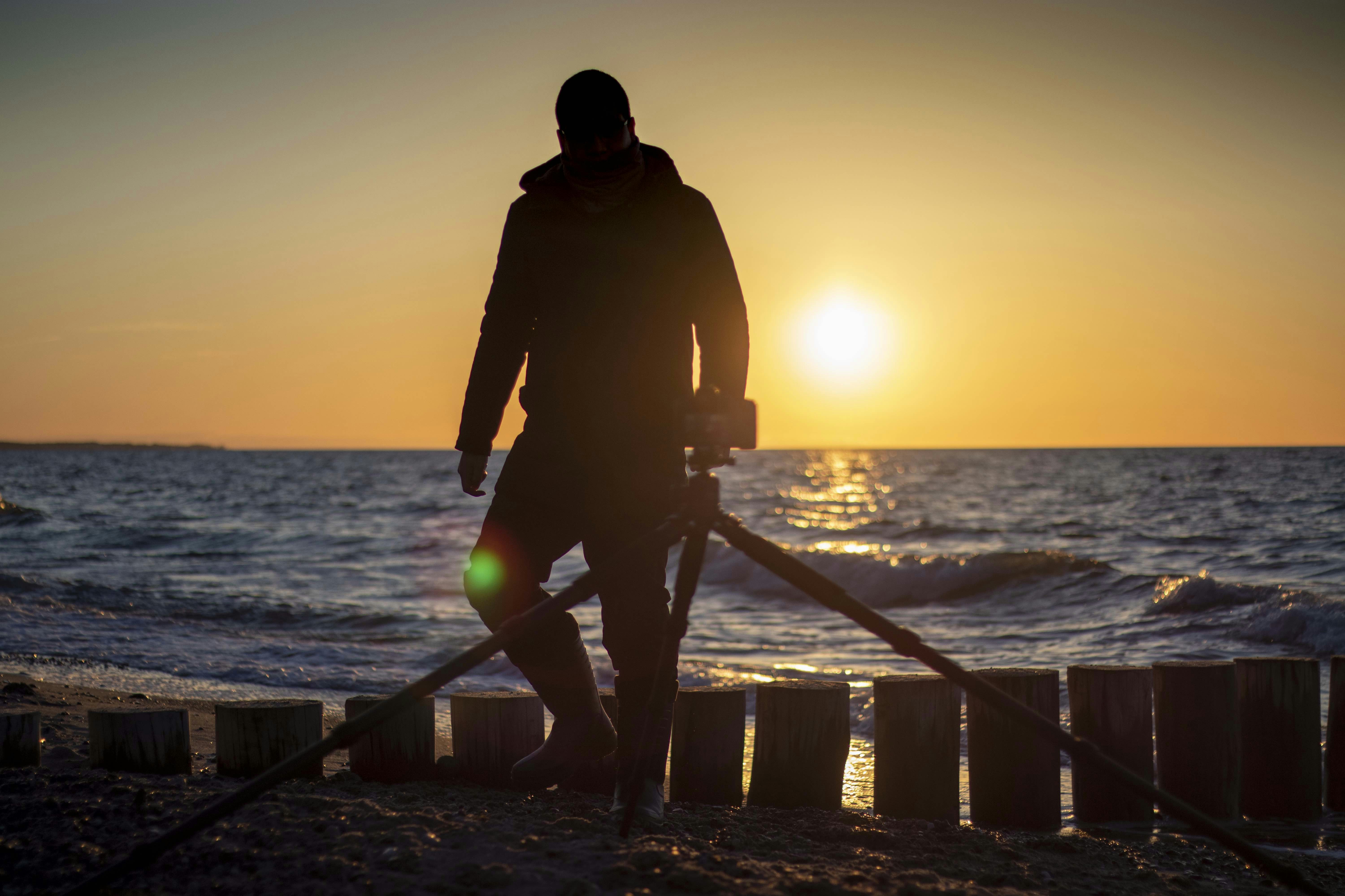Silhouette of a person standing on a wooden pier at sunset over the sea, the sun low on the horizon. The scene emphasizes mood and silhouette against warm sky hues.
