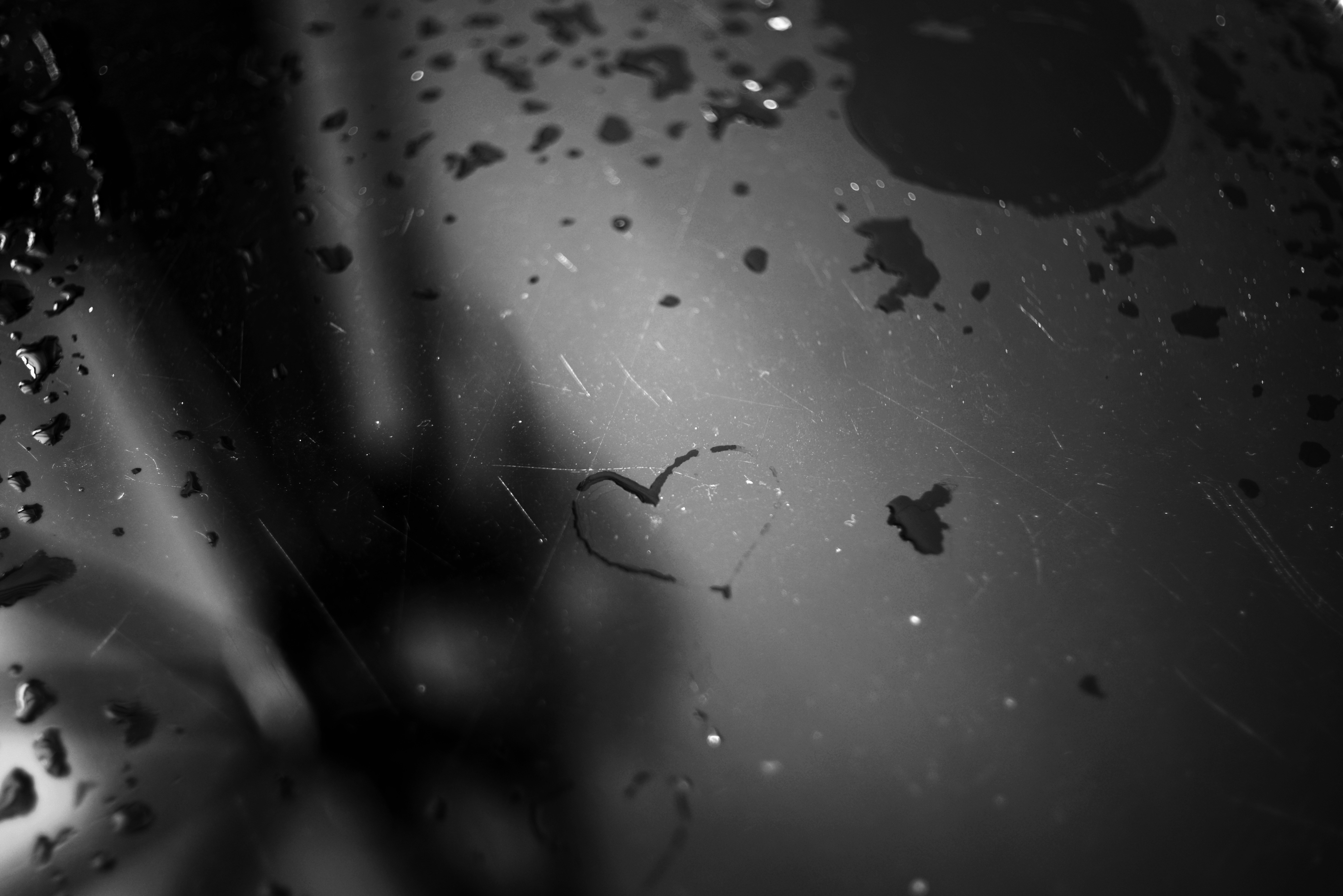 Monochrome close-up of a wet, reflective surface with scattered droplets and a dark silhouette in the background, emphasizing texture and abstract shapes.