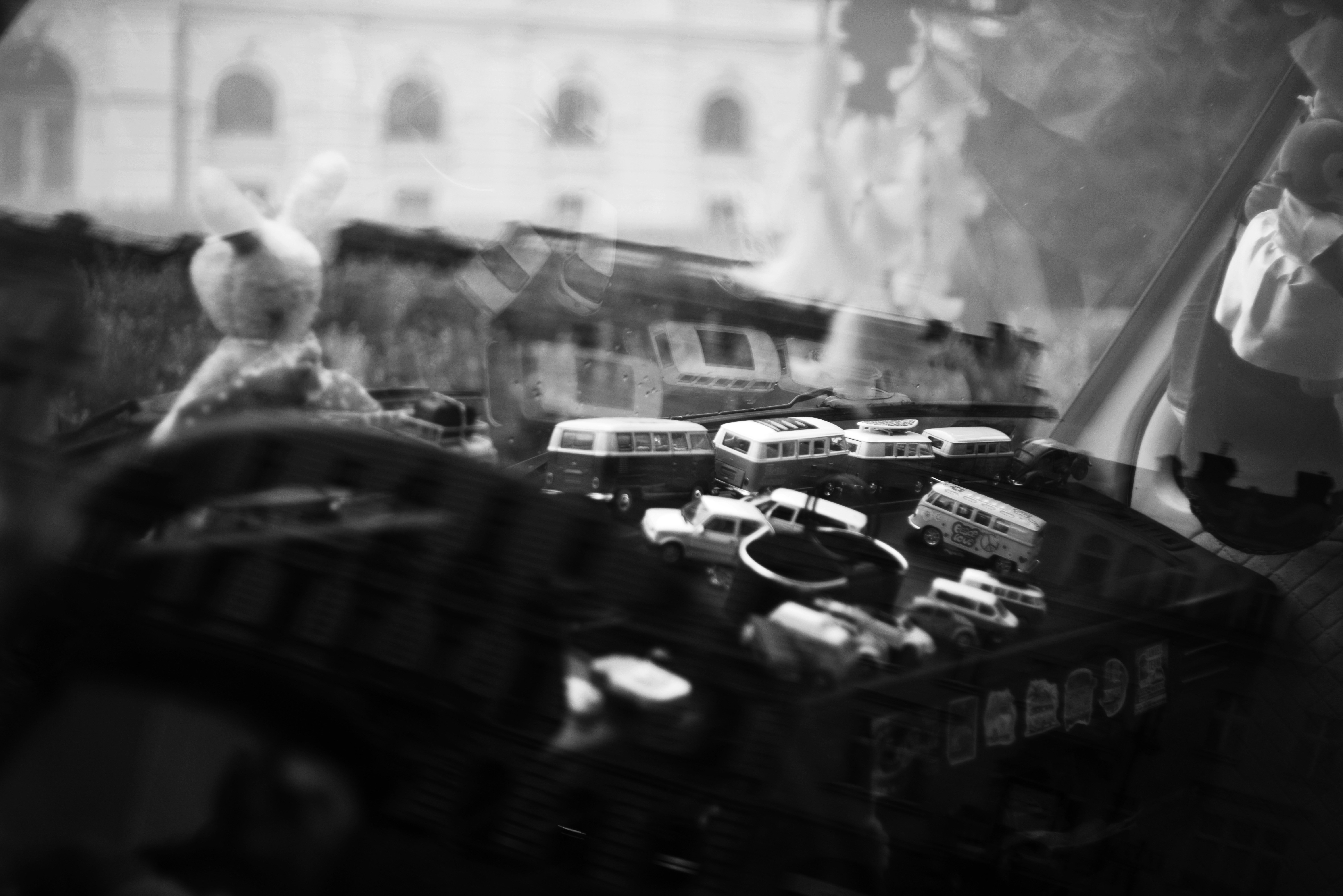 A grayscale scene of a cluttered surface with tiny toy vehicles and a reflected interior, blending toy displays with a window reflection.