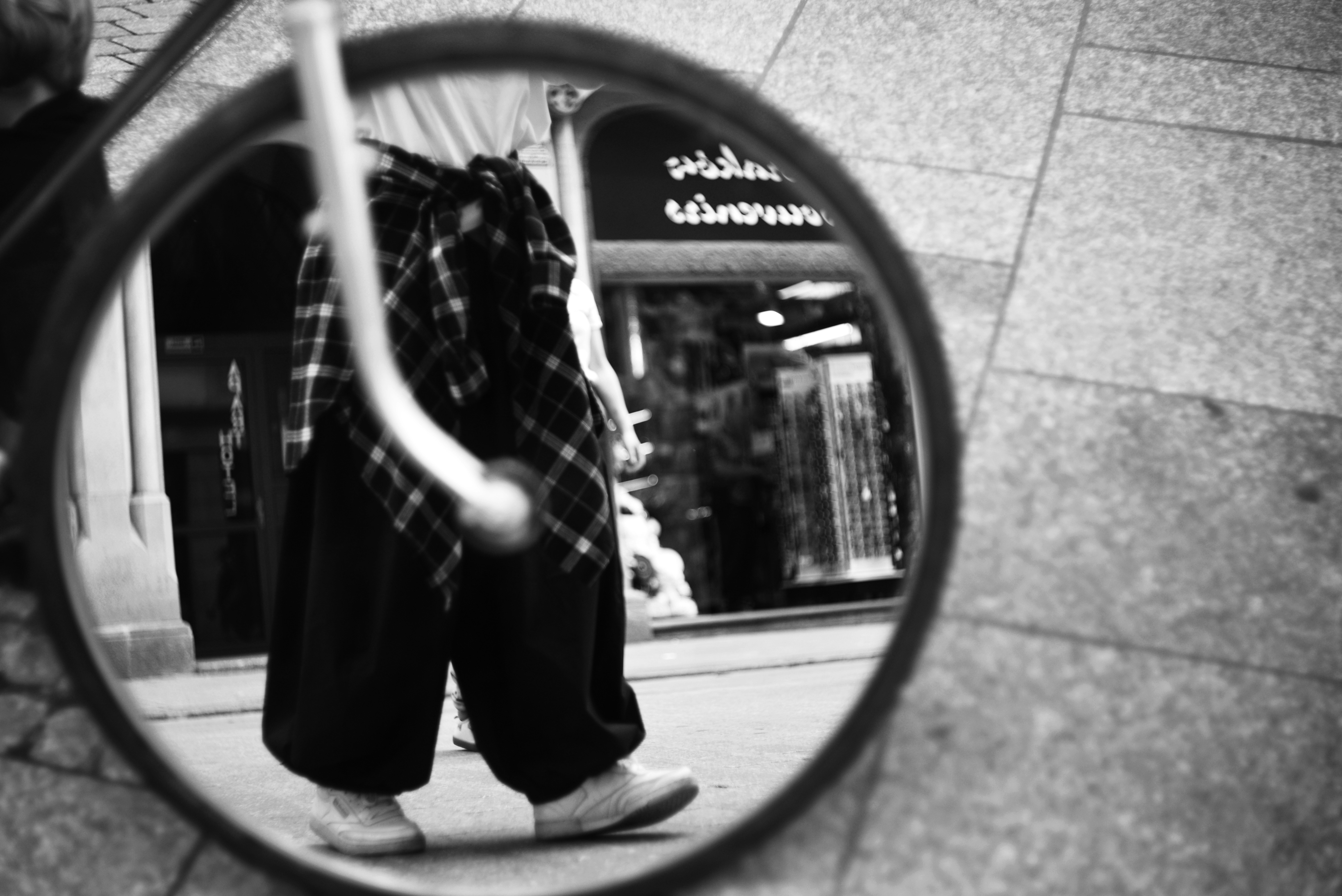 Street scene reflected in a circular window; a passerby’s legs and plaid shirt appear in the mirror, with surrounding architectural textures.