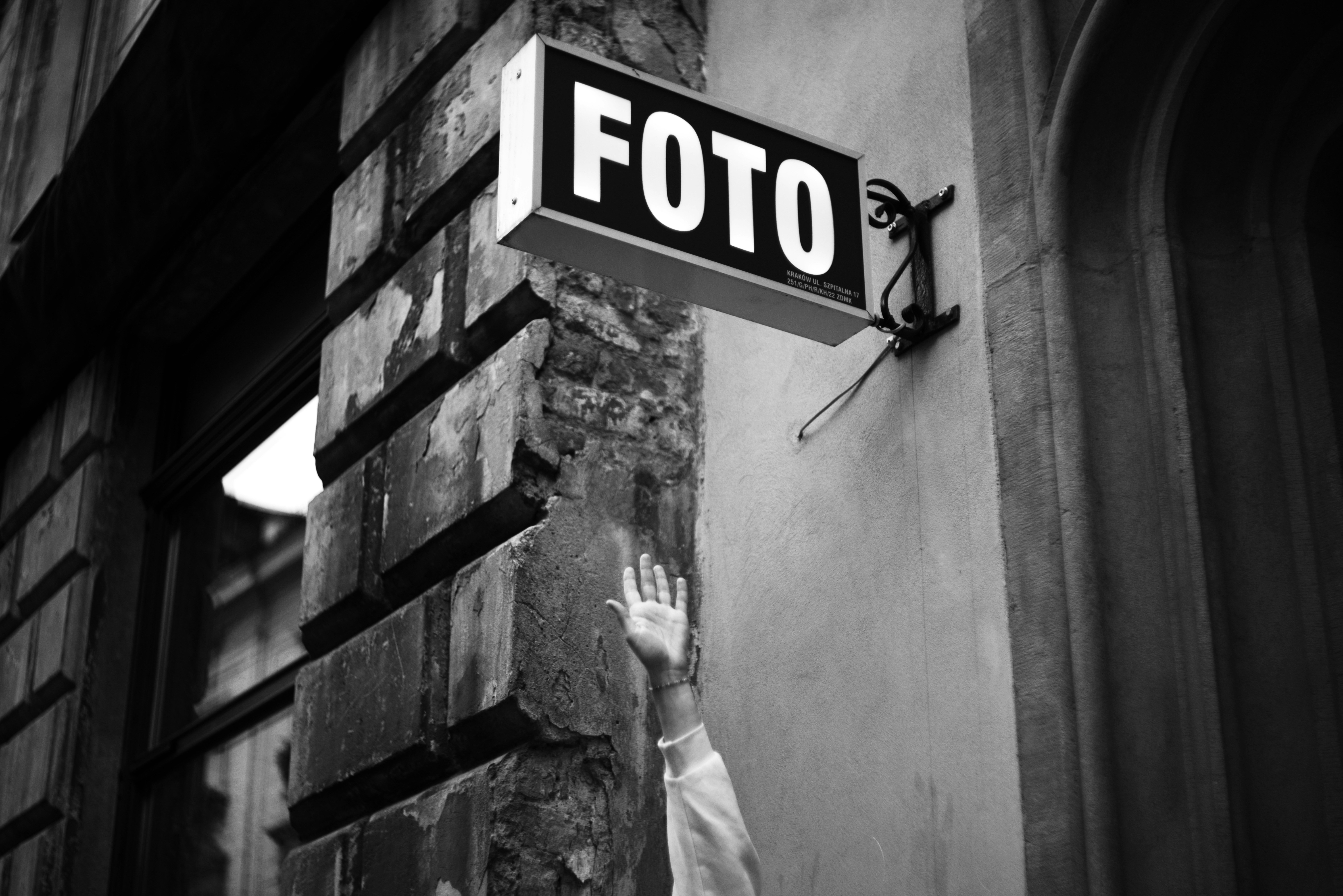 A grayscale street scene shows a solitary hand reaching up toward a bold FOTO sign above a rough stone facade. The image conveys a moment of access or attempt to enter a doorway, with architectural textures adding urban grit. Logo-like sign read as 'FOTO'.