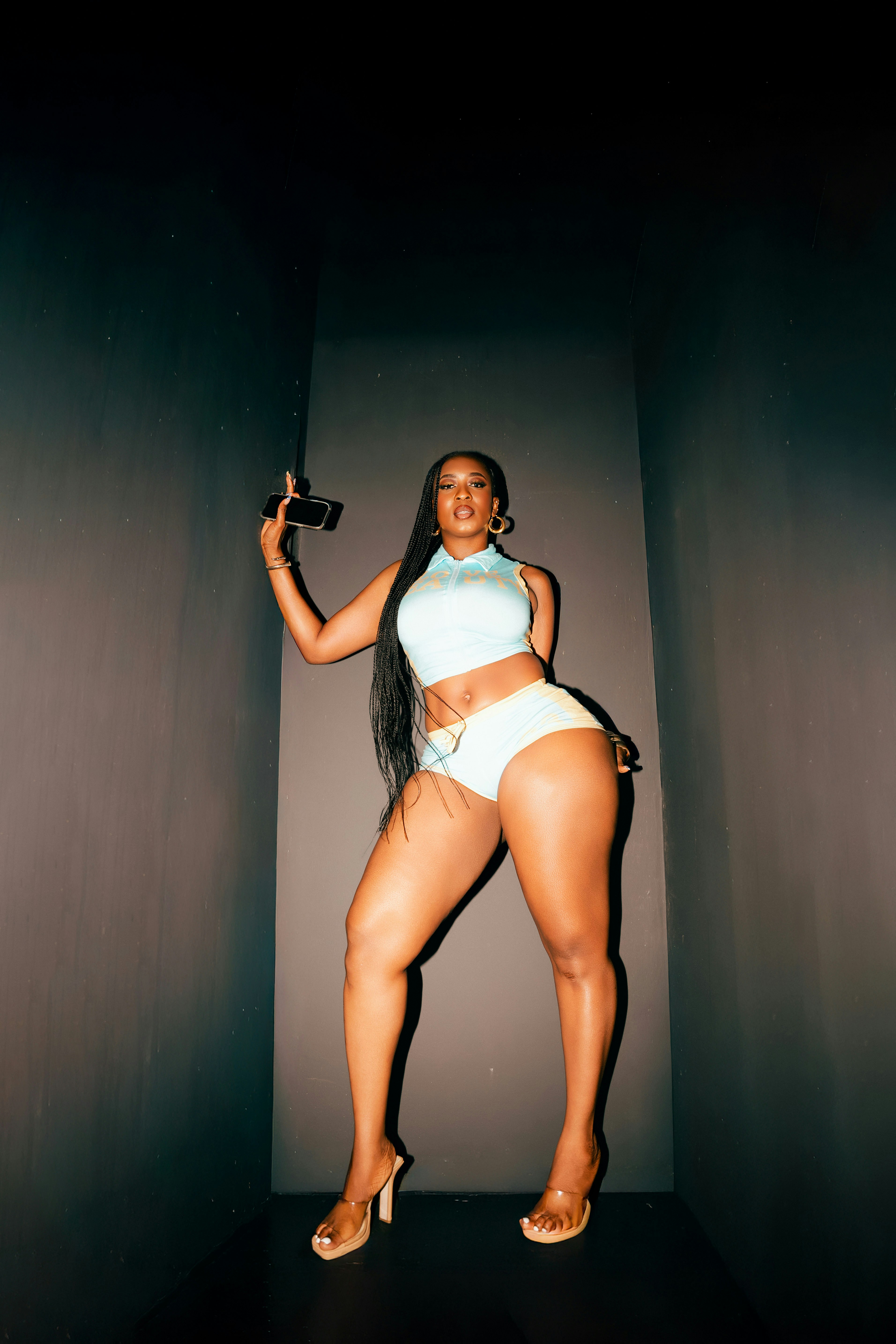 Slim silhouette stands center stage in a tight, pale-blue outfit, posed with a phone in hand inside a narrow, dark alcove. A bold, fashion-forward portrait for a club or event setting.