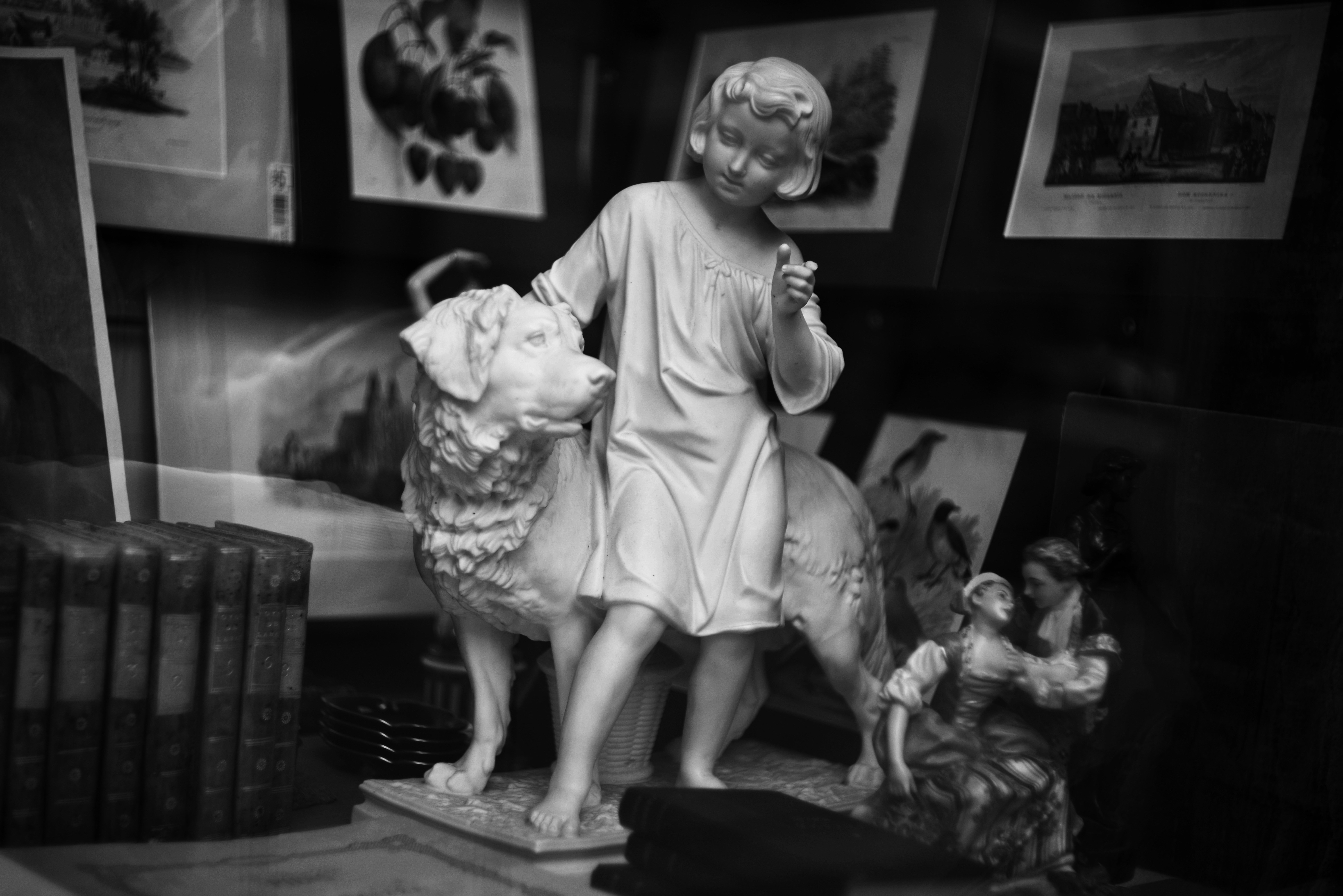 A grayscale display scene featuring a porcelain child statue with a sheep, surrounded by framed pictures and books behind glass.