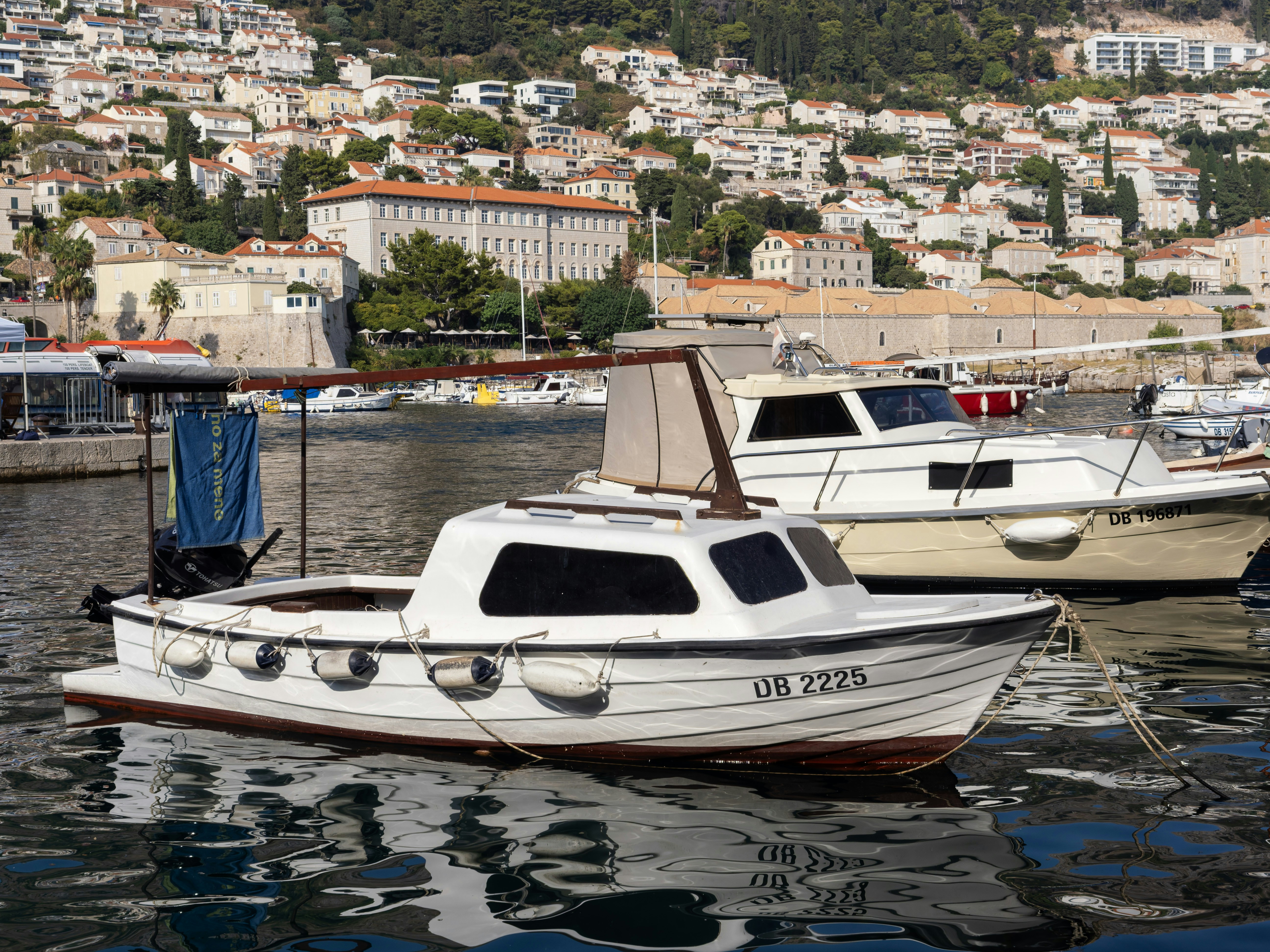 A white motor boat rests on calm harbor waters with a bustling town and hillside homes in the background, conveying a tranquil coastal scene.