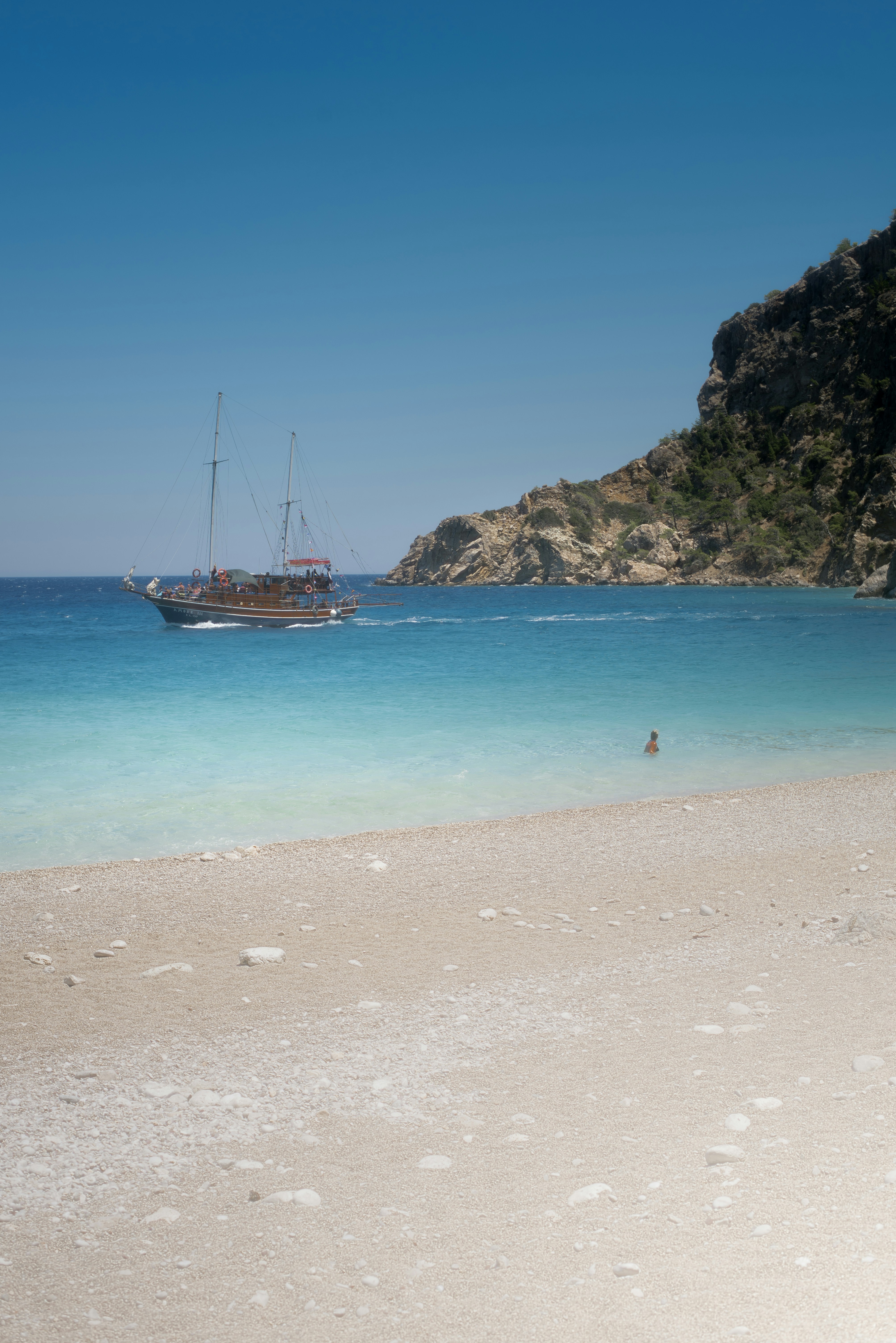 Caribbean-like turquoise water kisses a pebble beach with a classic sailboat anchored near a rocky headland.