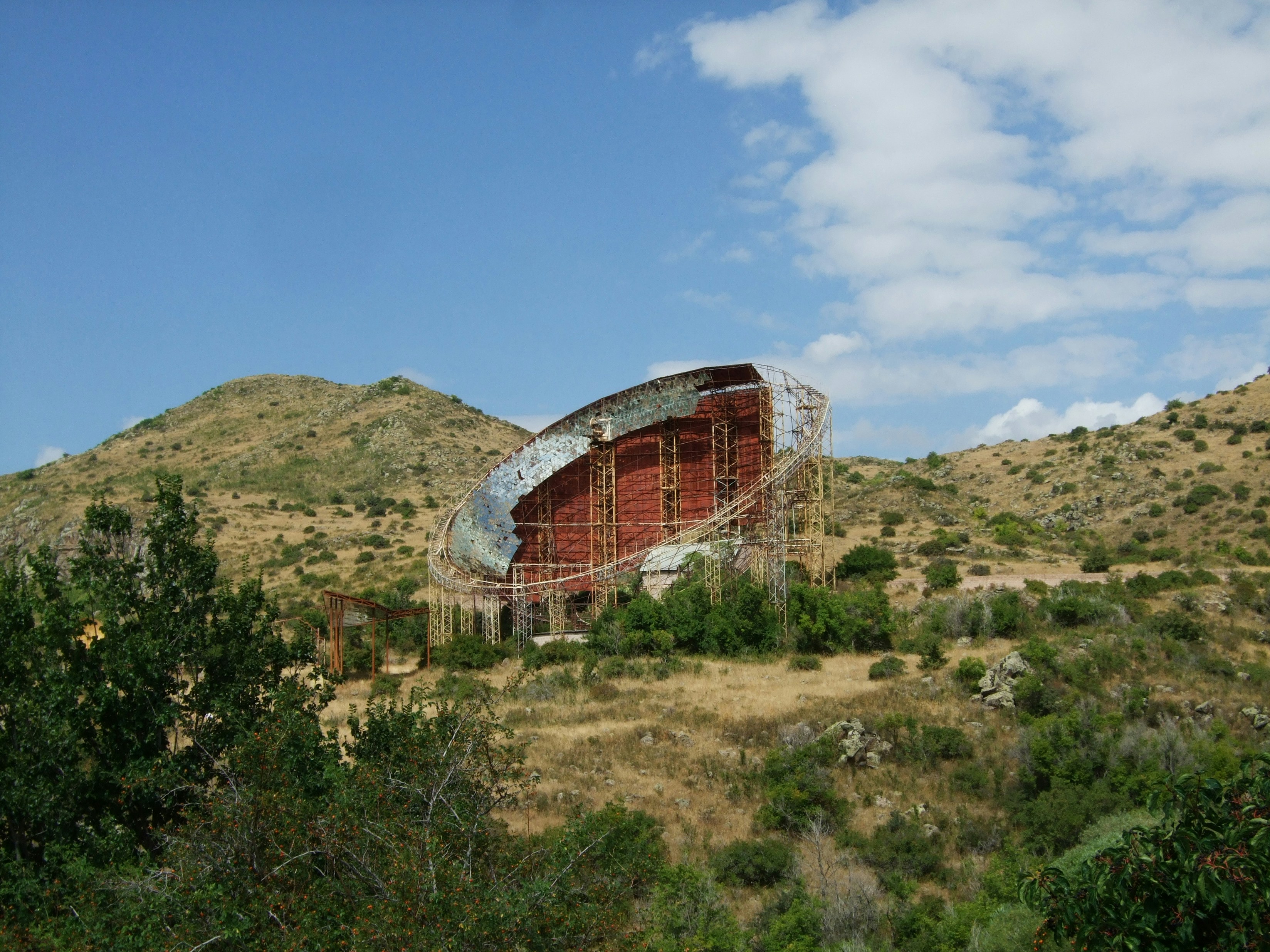 A large rusted parabolic antenna structure on a dry grassy hillside under blue sky