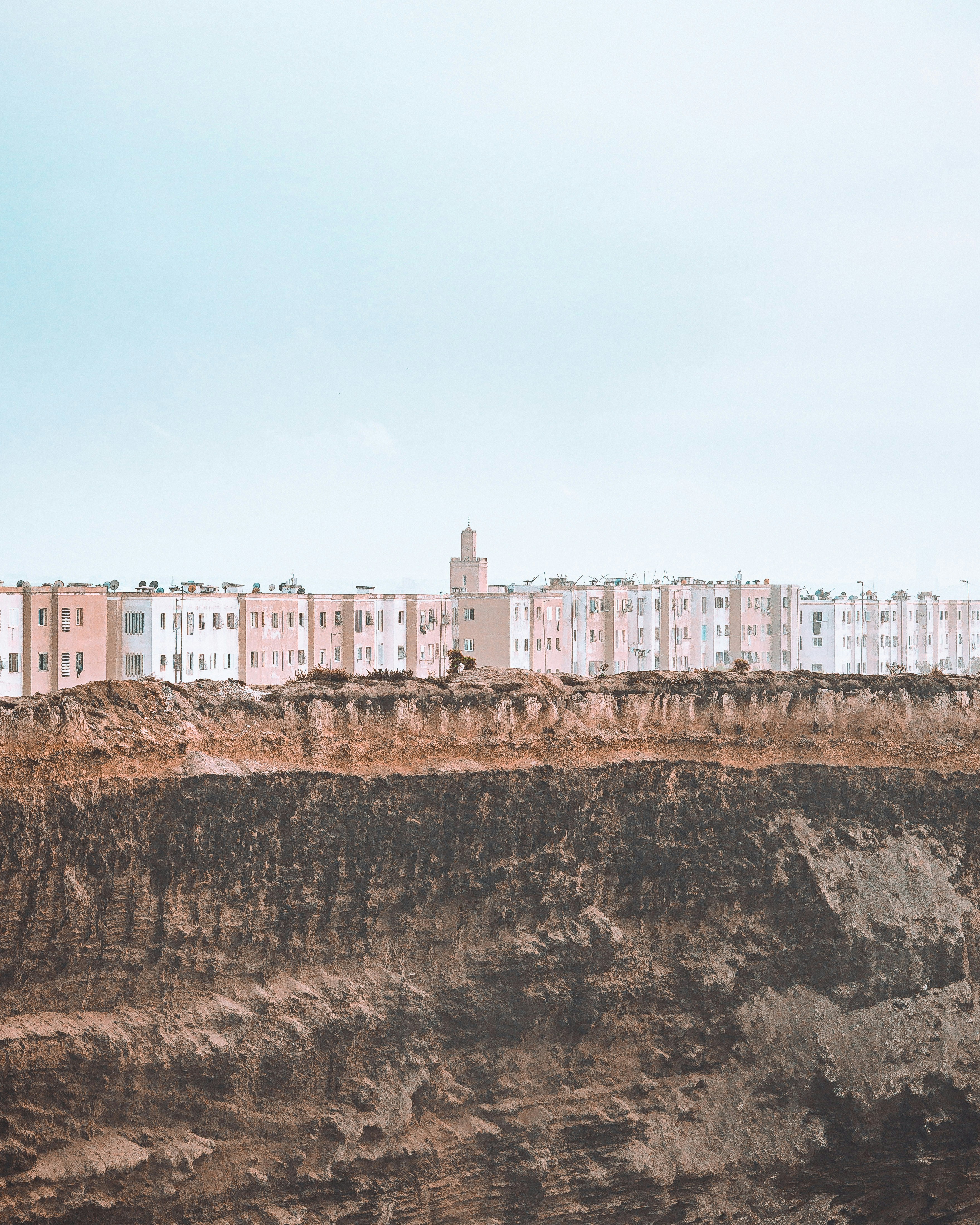 Row of pastel apartment buildings perched along a cliff edge with a clear sky above; the scene emphasizes layered geology meeting urban housing.