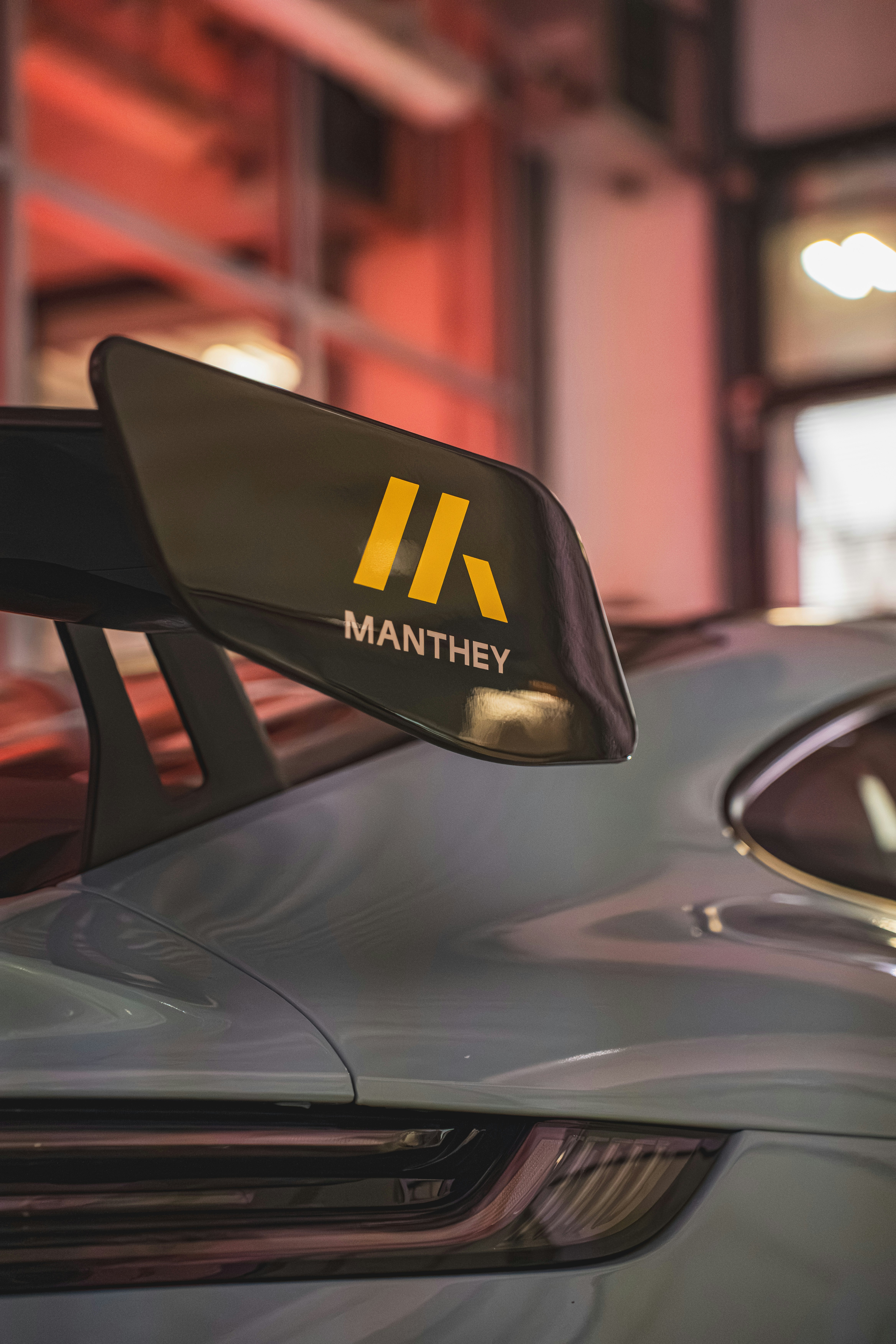 Close-up of a light blue sports car with a dark rear wing bearing Manthey branding in a softly lit urban setting. The focus is on the spoiler and rear window area, with blurred background.