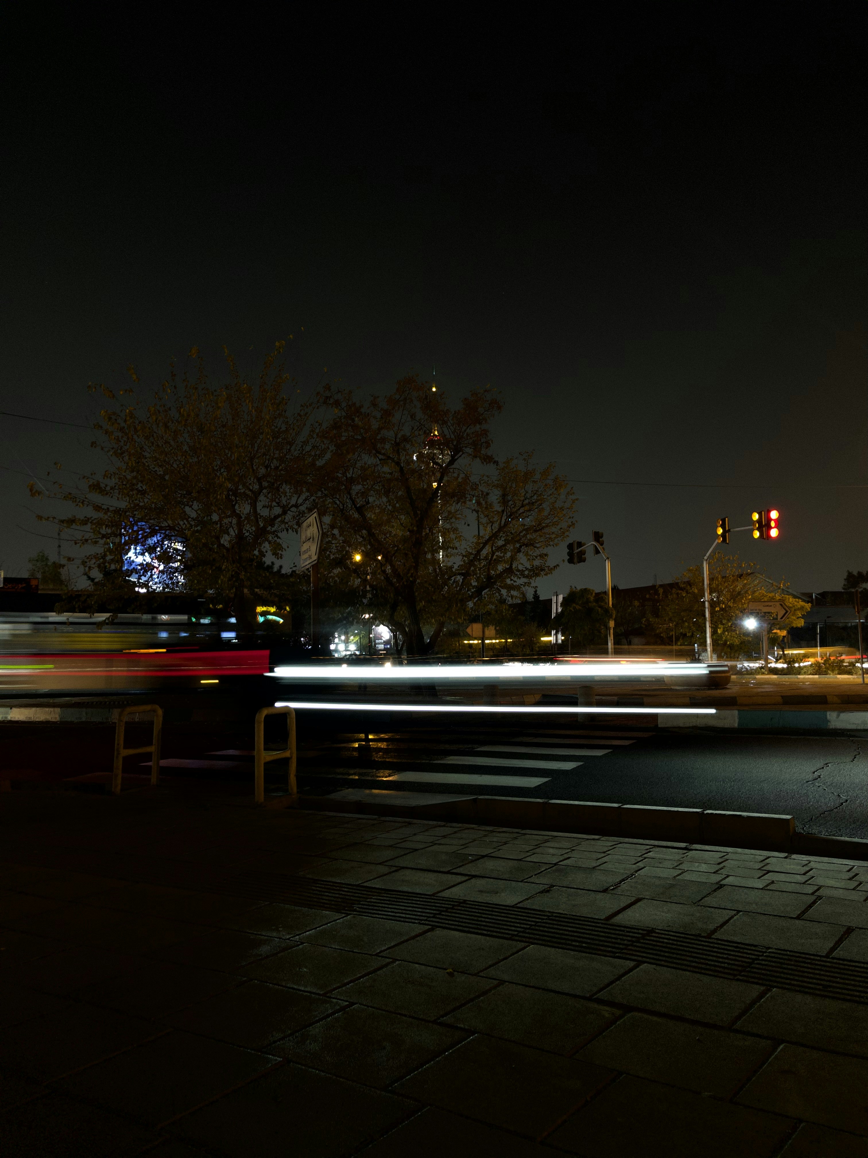 Nighttime street scene with blurred vehicle light trails and a dark sky; foreground shows tiled pavement and street furniture, while distant city lights and a tall building silhouette create depth.