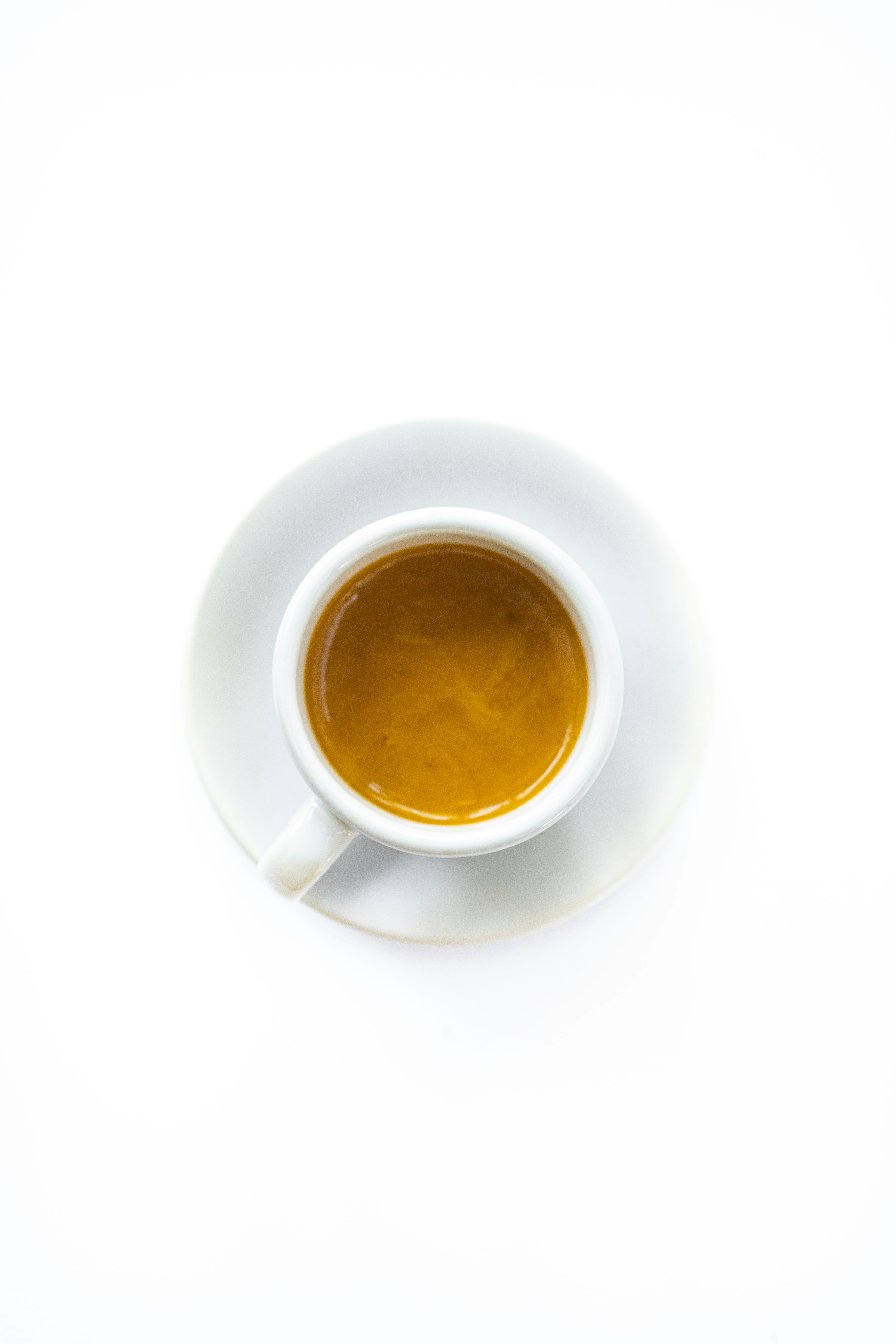 Top-down shot of a white porcelain cup of espresso on a white saucer, centered with soft shadows and minimal background; a simple still-life emphasizing coffee's amber tones. This is a photography capture, not a drawing or painting.