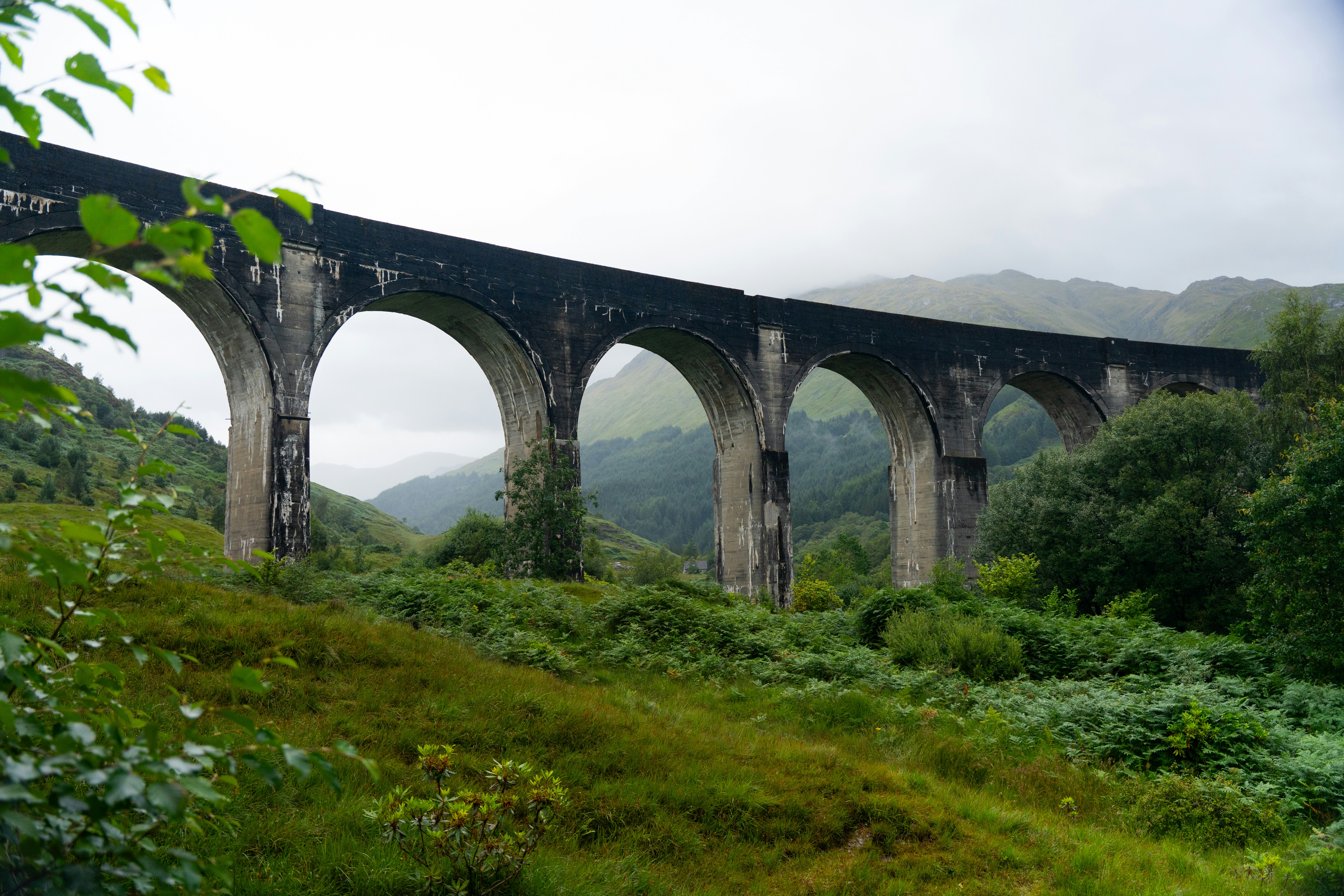 Stone arches of a historic viaduct rise over lush green hillside with distant misty mountains, set under a pale sky; an outdoor architectural scene captured in wide landscape.
