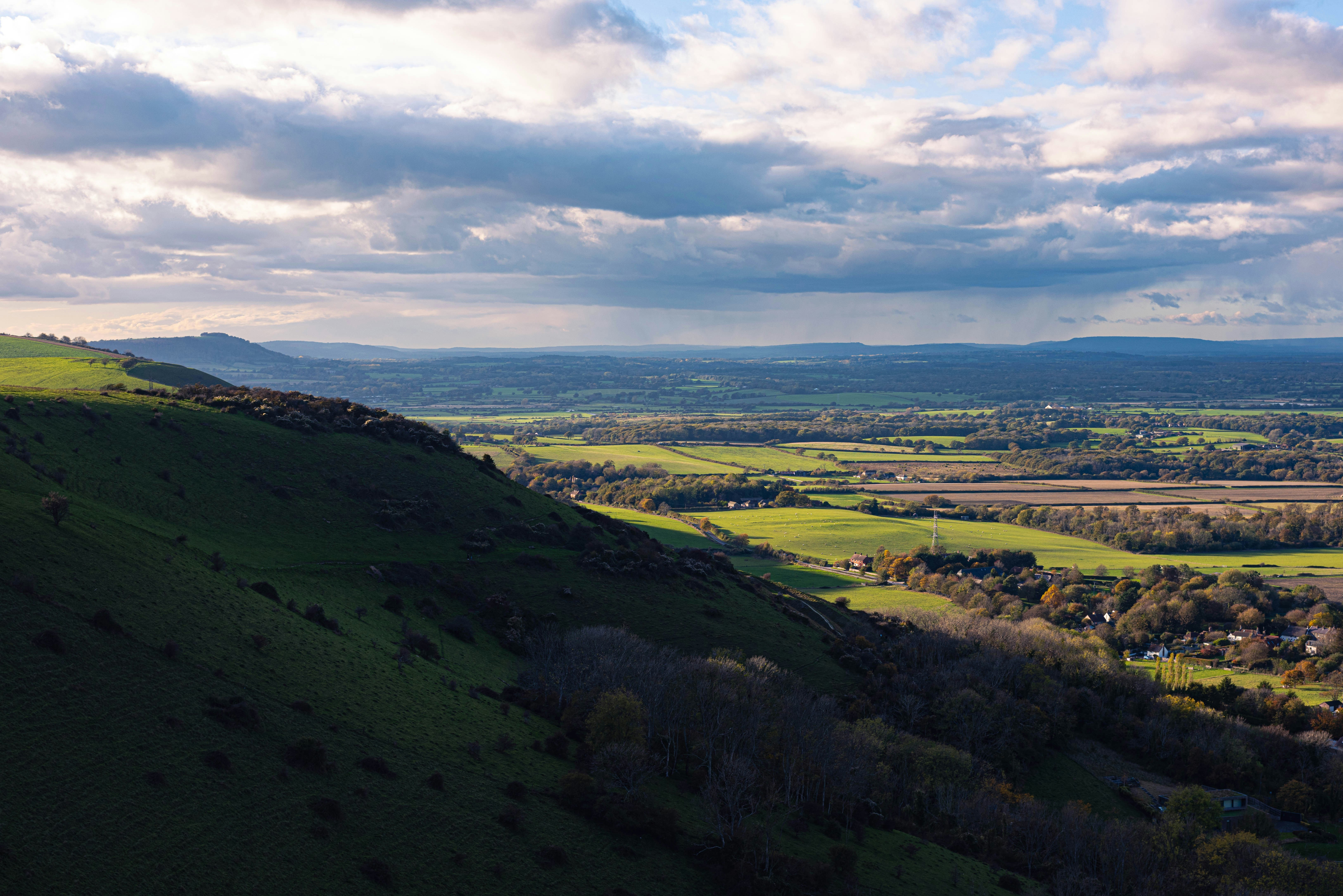 Vast landscape with rolling hills, patchwork fields, and a distant horizon under a dramatic sky; a subtle contrast between sunlit valleys and shaded hillside.