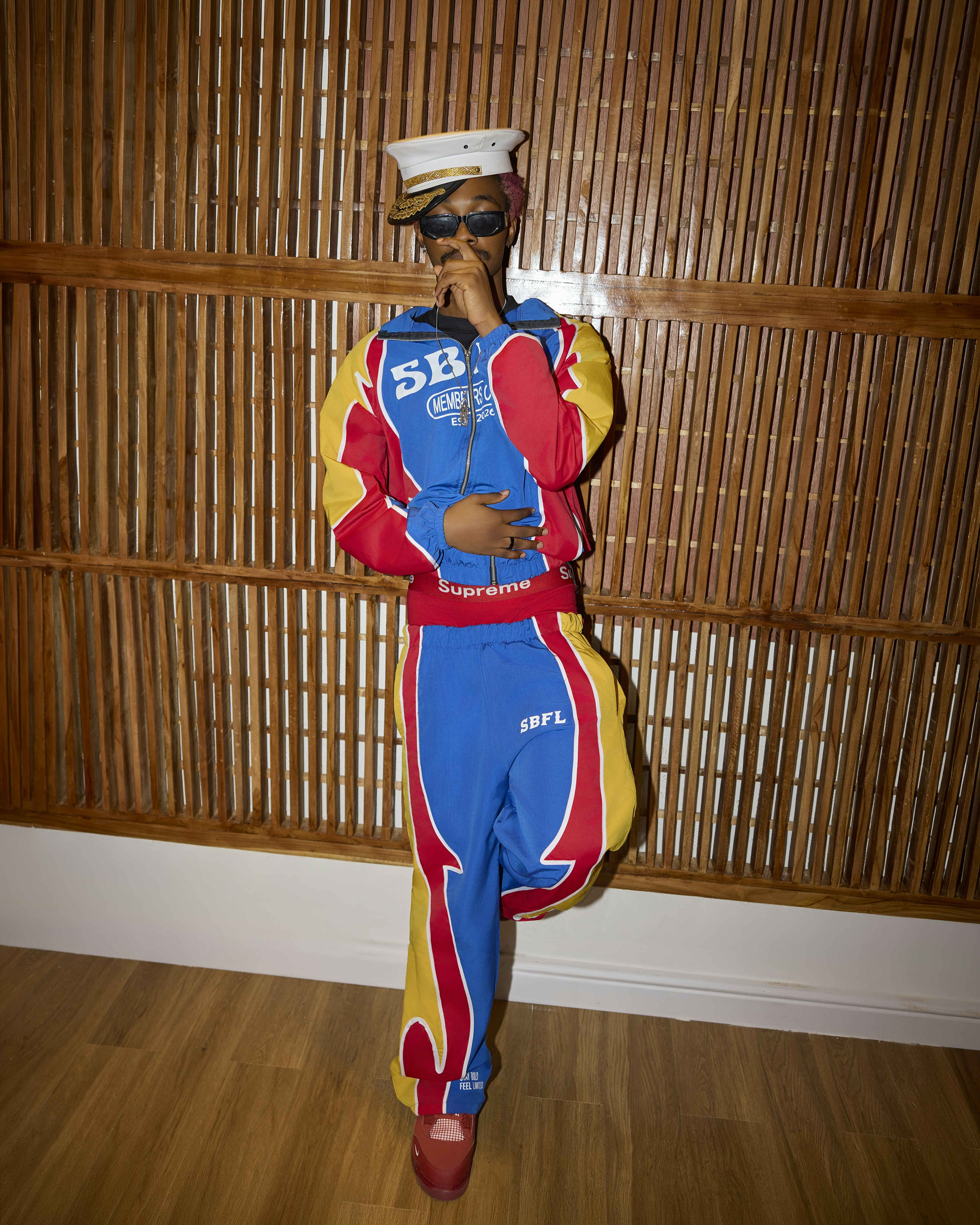 Portrait of a person leaning against a wooden slat wall, wearing a blue-red-yellow color-blocked tracksuit and a white captain’s hat, with sunglasses and a confident pose. A logoed, sporty outfit dominates the frame.