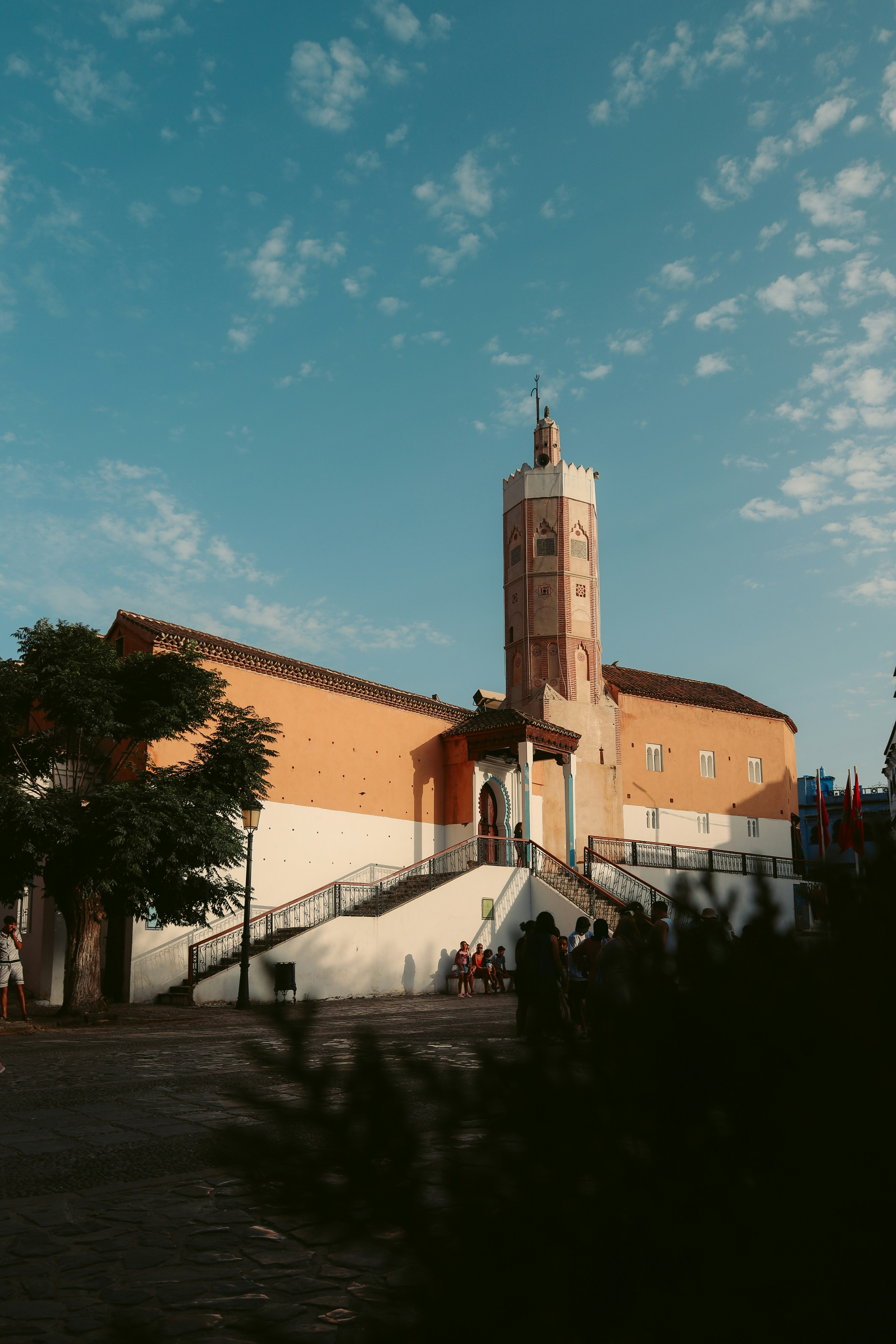 Sunlit fortress with a tall central tower and orange walls sits under a clear blue sky; a small crowd gathers at the stairs leading to the entrance (logo/crest visible in the façade).