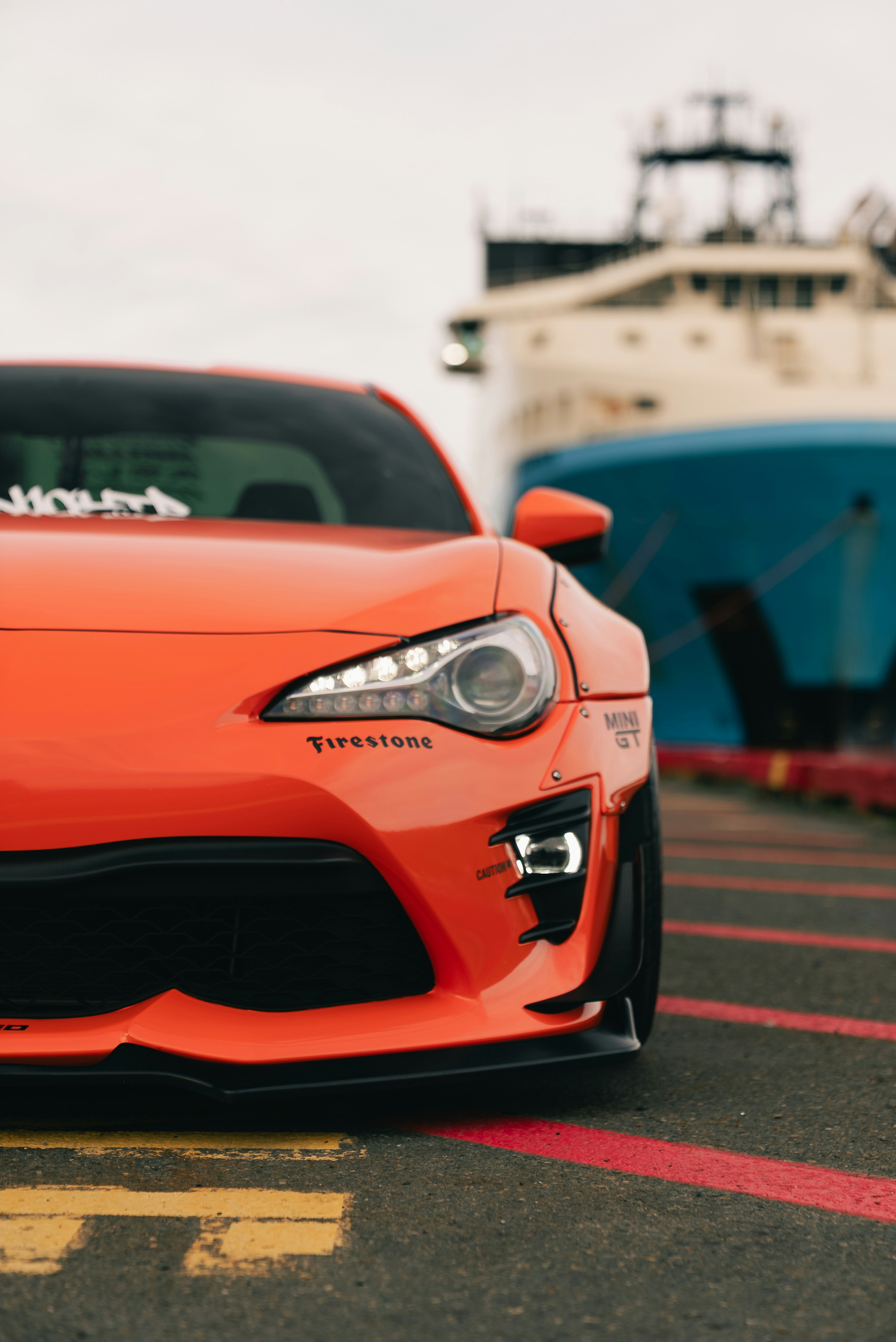 Fiery orange sports car parked on a dock, front-left corner framing the vehicle with a blue ship in the background; a crisp automotive detail shot suitable for a dealership or editorial use.