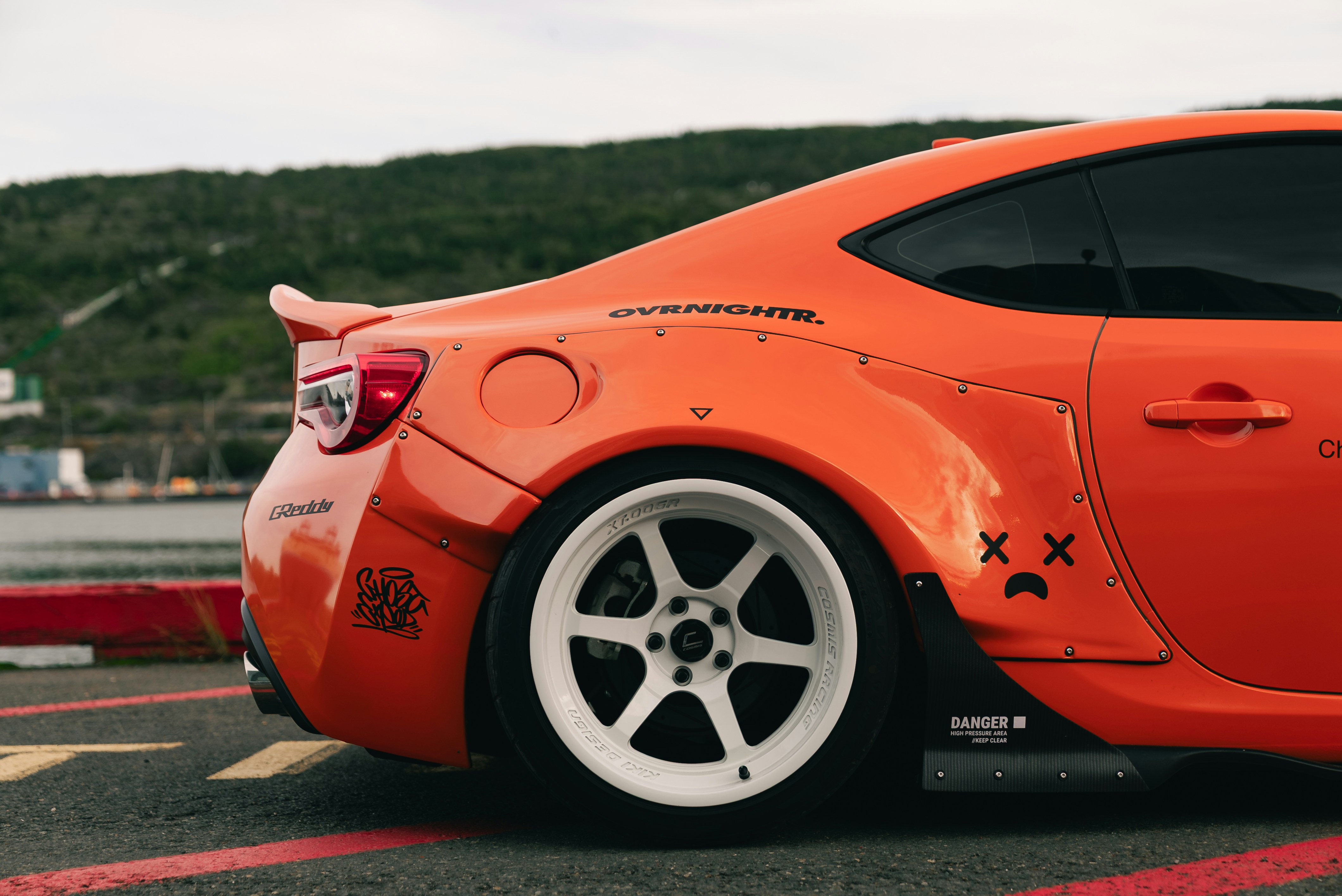 Bright orange sports car with white wheel, shown from a low angle on a racetrack; emphasis on rear quarter panel, tire, and decals. Logo/graphics present on body in a parking-lot setting.