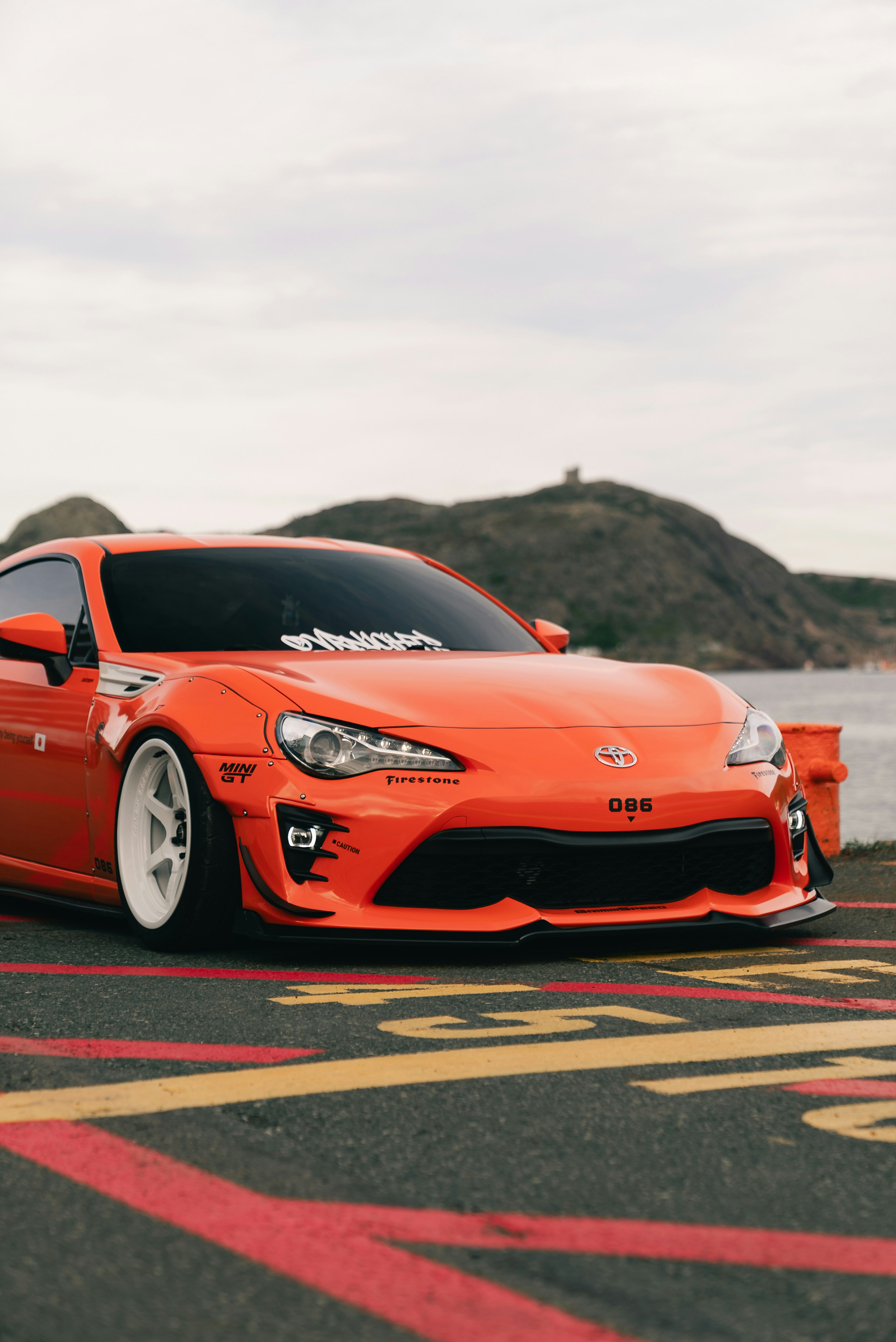 Vibrant orange sports car parked on a marked surface with distant rocky hills under a soft sky.