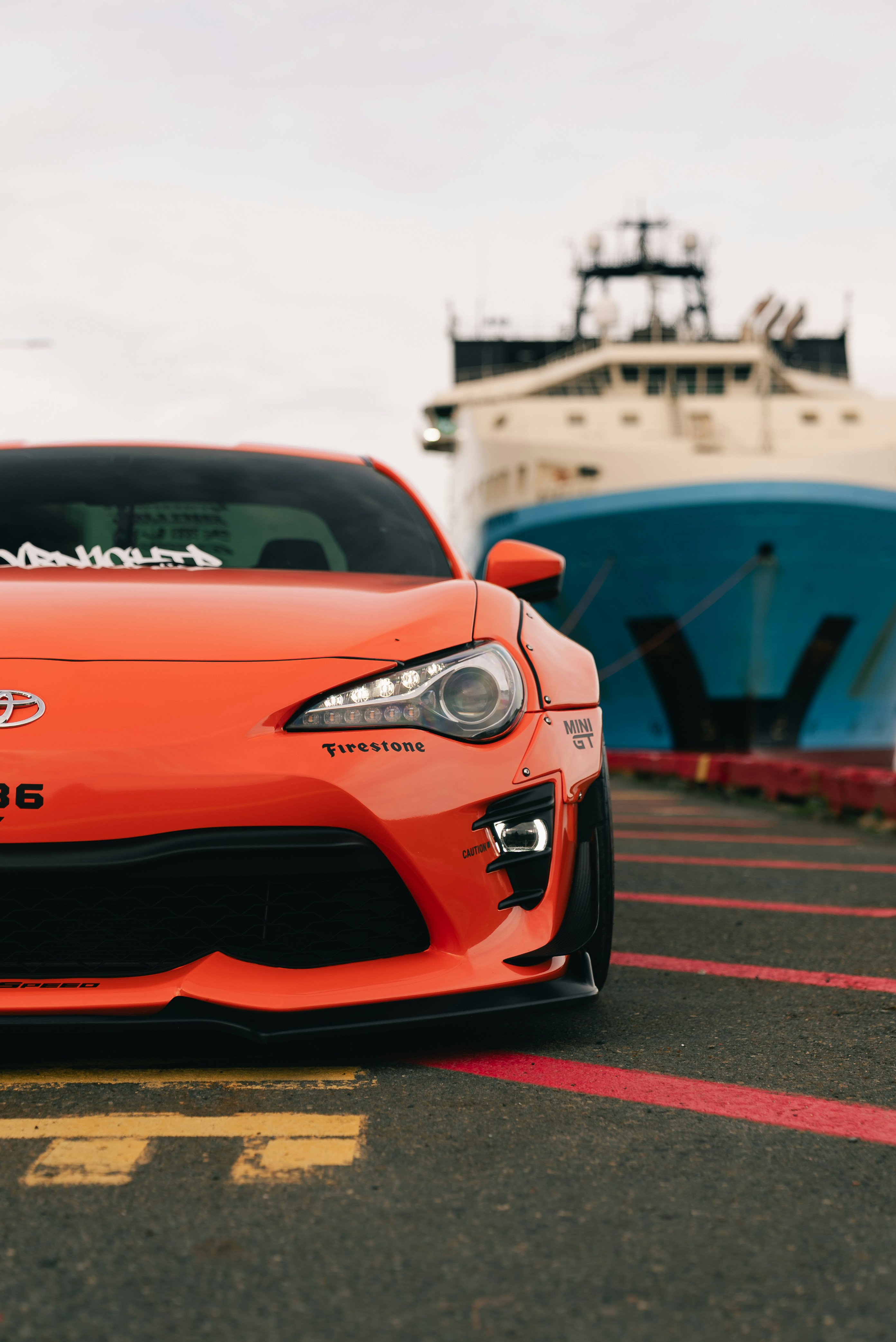 A bright orange sports car parked at a dock, with a large blue ship in the background. The low-angle composition emphasizes the car’s curves and branding.
