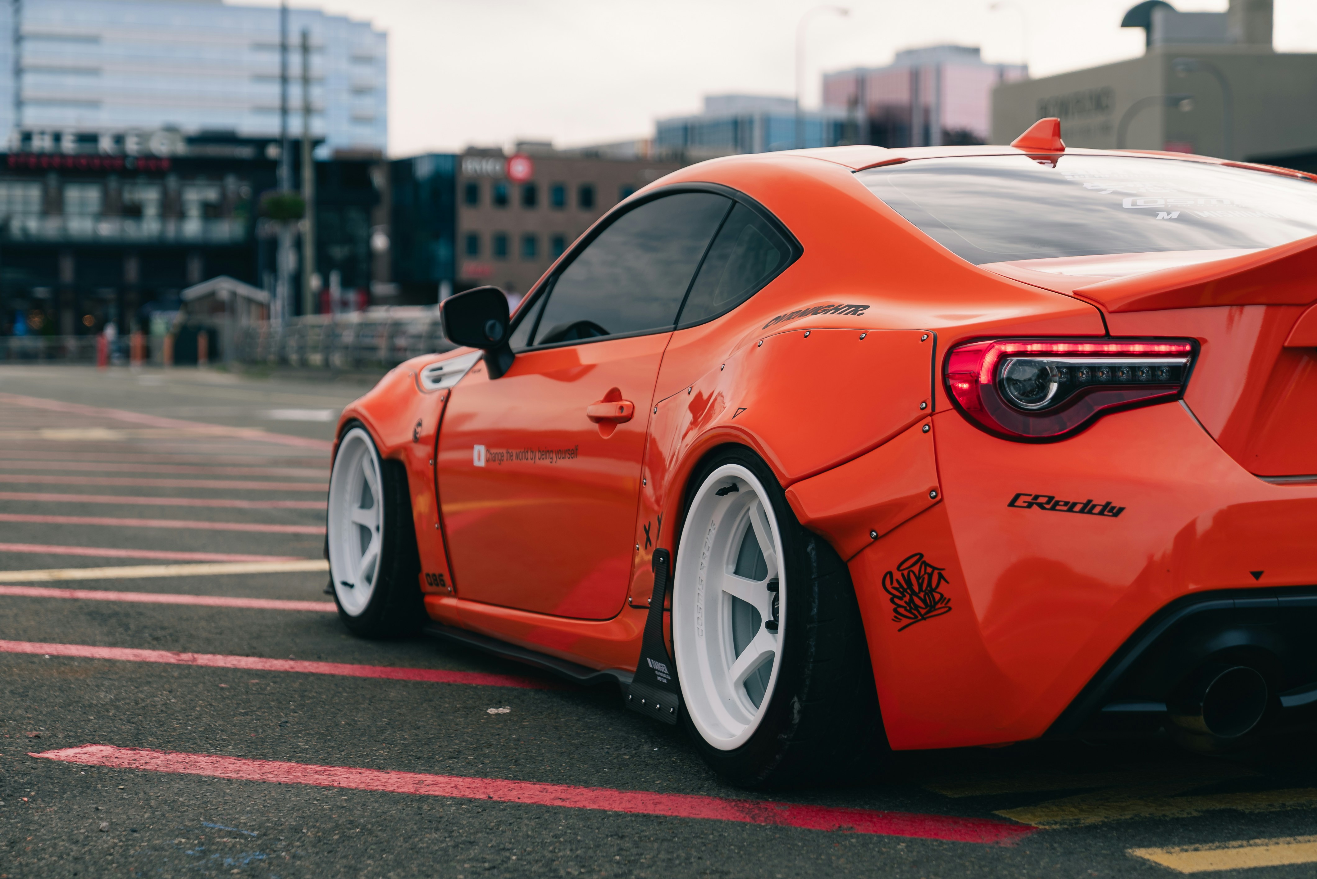 Orange sports car with wide white wheels parked in a lot, rear three-quarter view showing badge and decals. A muted urban backdrop emphasizes the vehicle's stance and details.