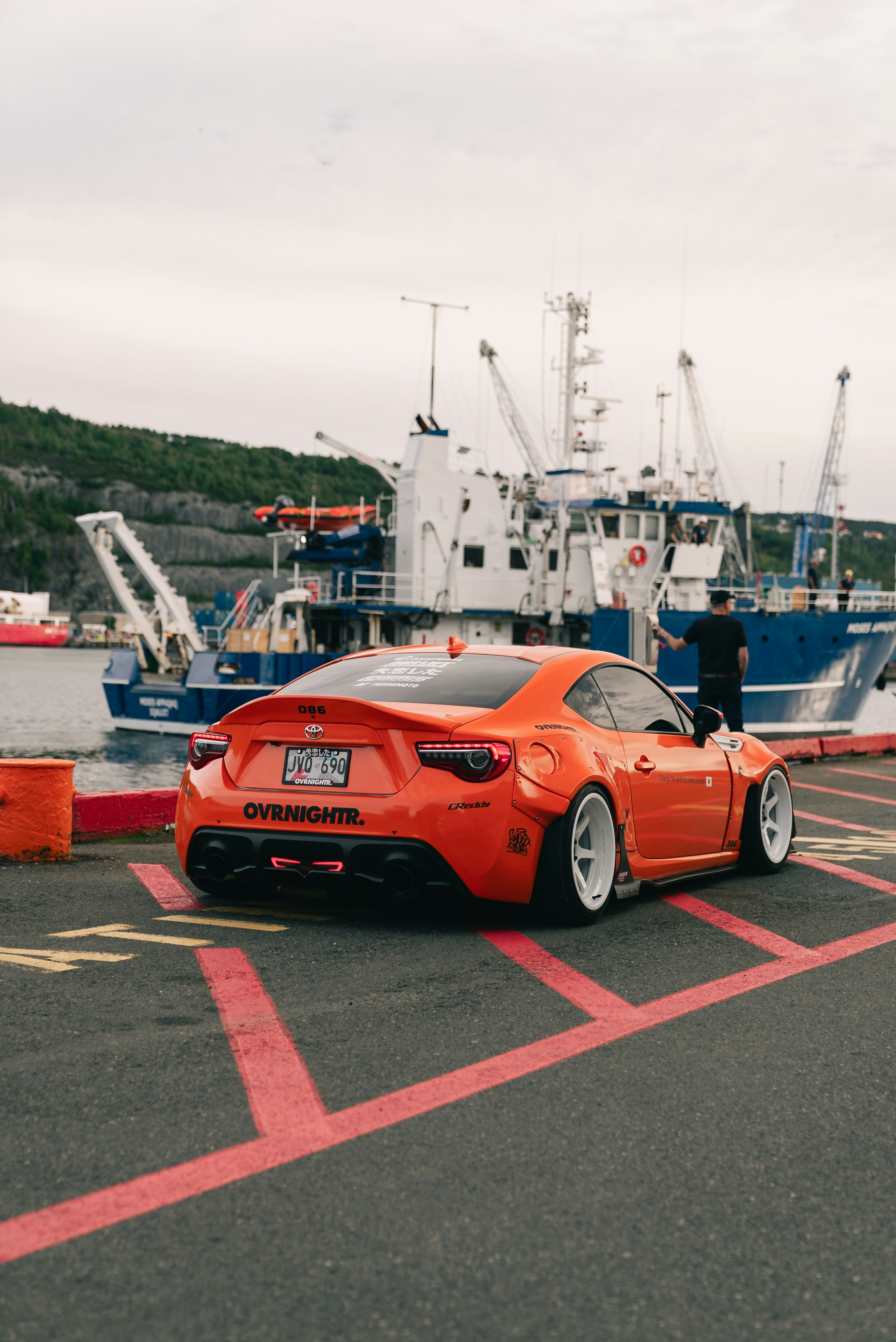 Bright orange sports car parked at the waterfront near cranes and a blue ship; foreground lines mark a red grid on the pavement. This is a photograph capturing a car scene at a harbor setting.