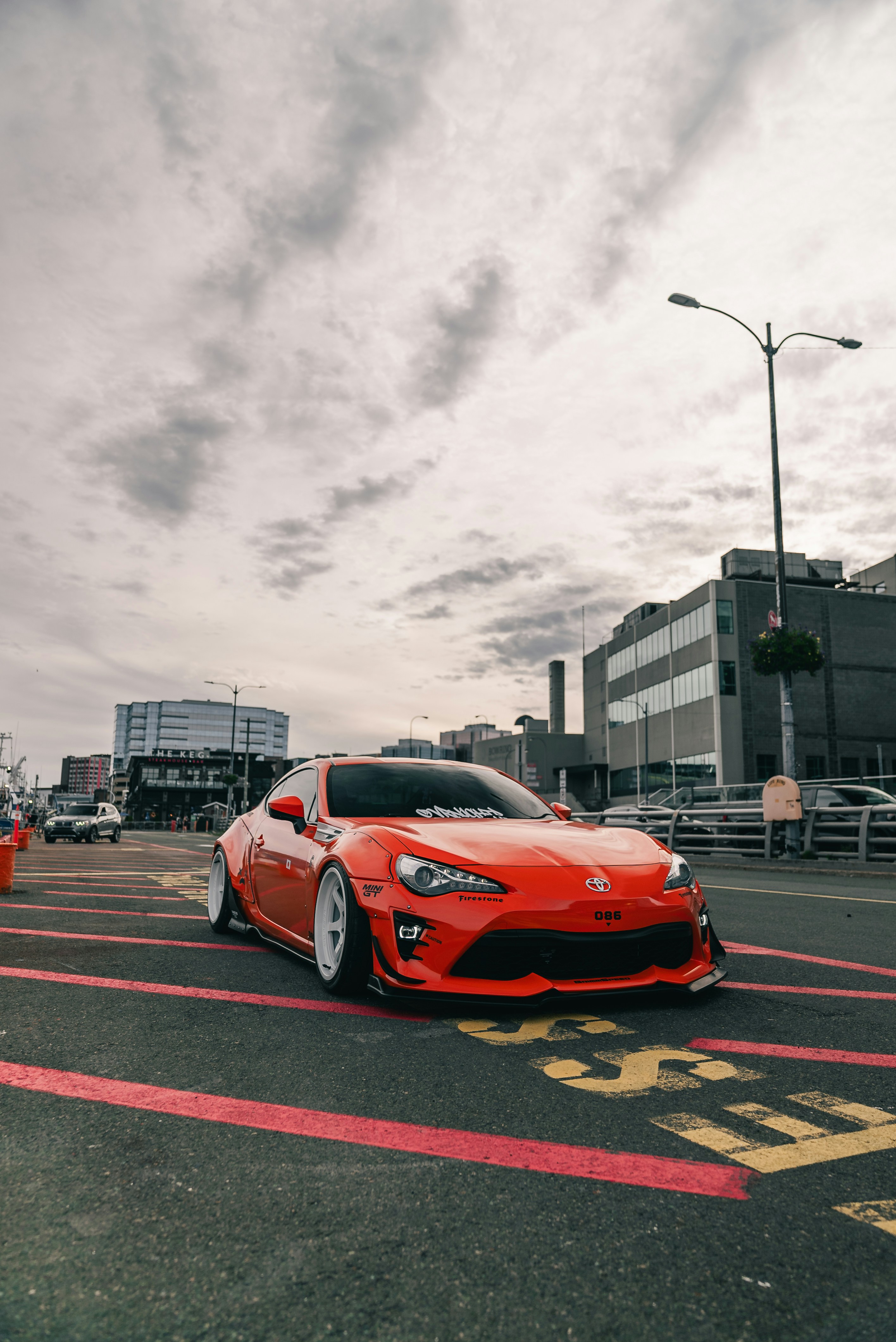 Red sports car parked in a marked lot with an urban backdrop of industrial buildings and a cloudy sky, emphasizing low-angle perspective and bold color. This is a photographic capture likely shot in a parking area.